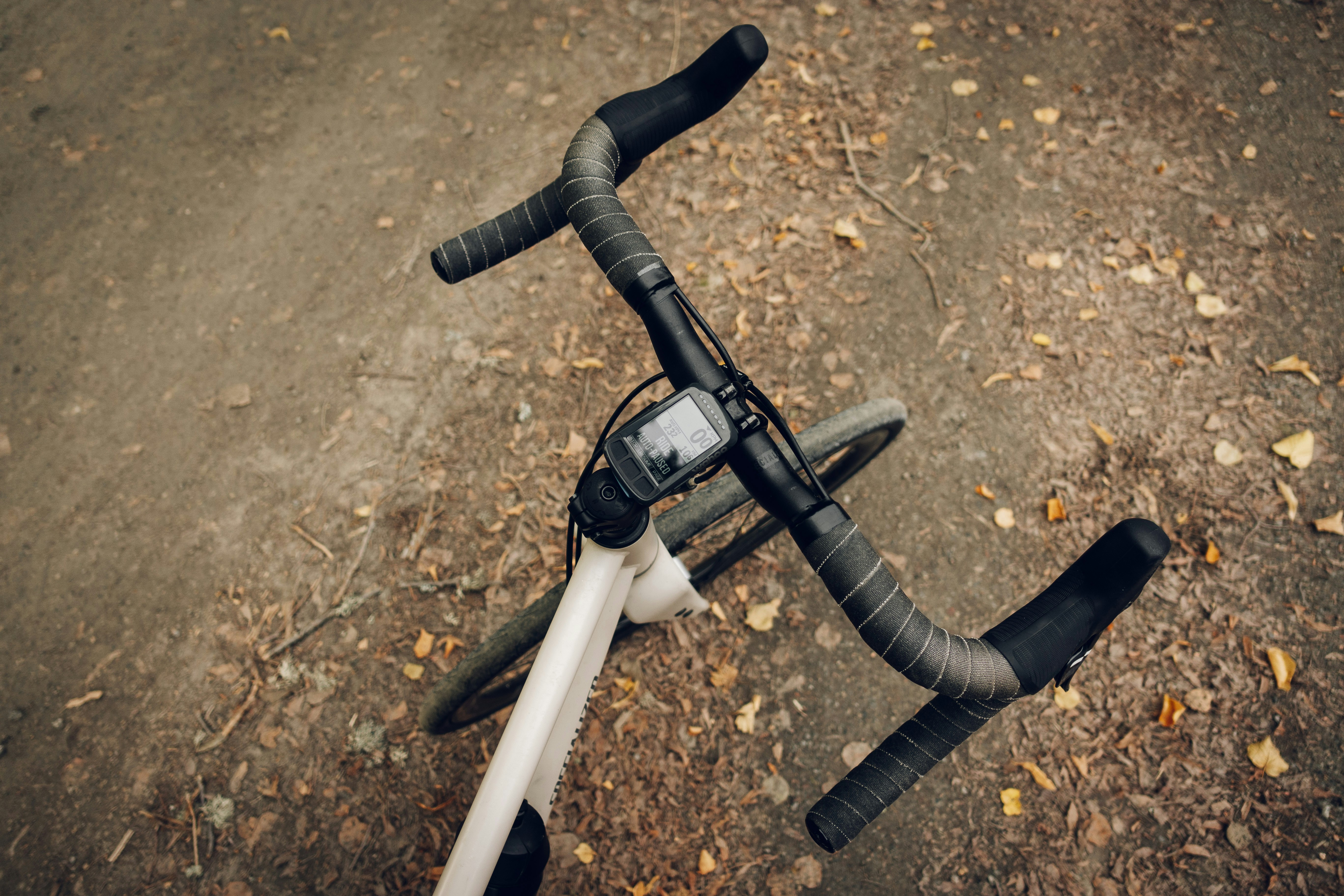 Overhead view of a bicycle's handlebars resting on a dirt path, showcasing the textured grips and cycling computer. The ground is scattered with leaves, hinting at an autumn ride.