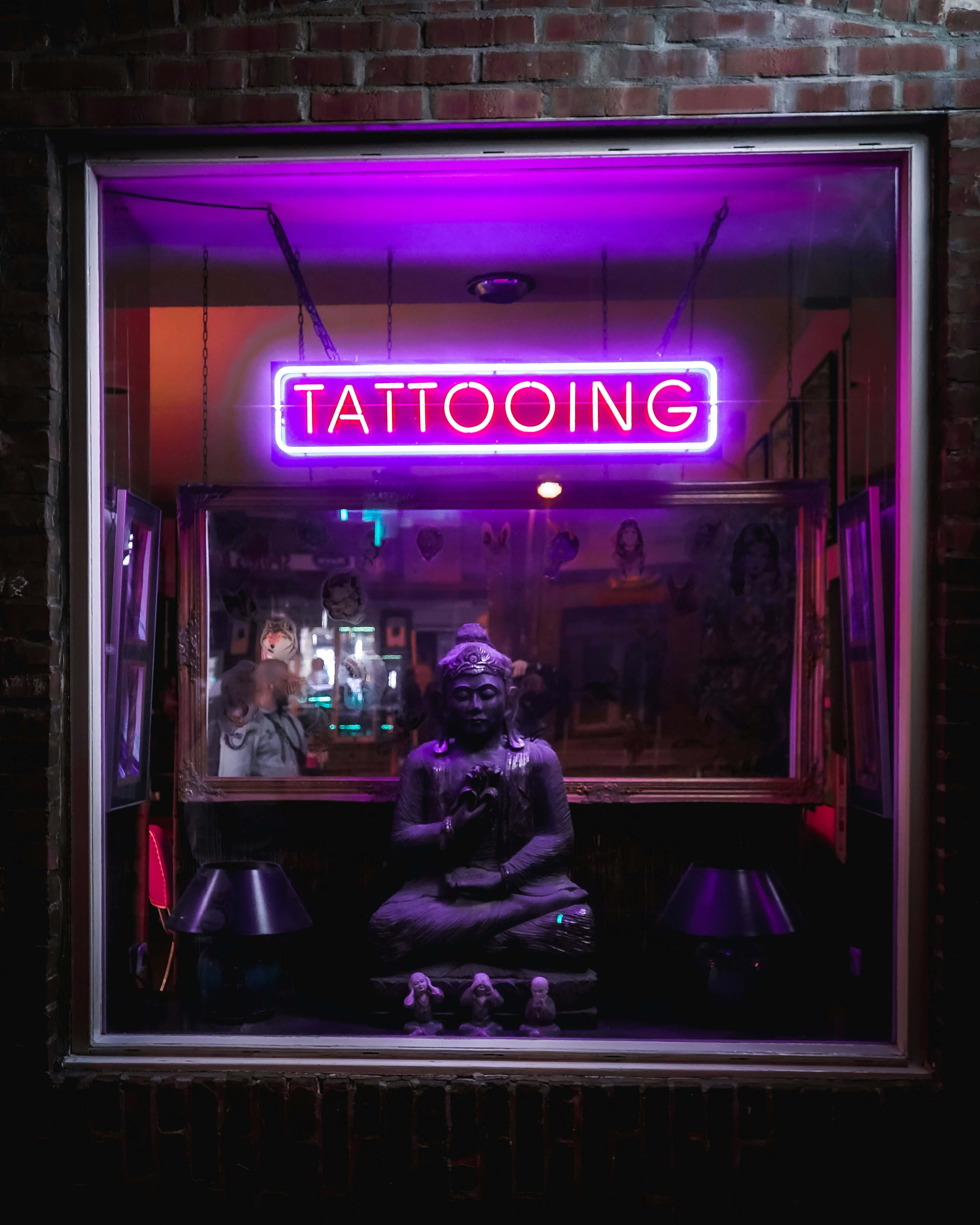A serene Buddha statue sits in a tattoo parlor, framed by a vibrant neon 'TATTOOING' sign. The warm glow creates an inviting atmosphere.