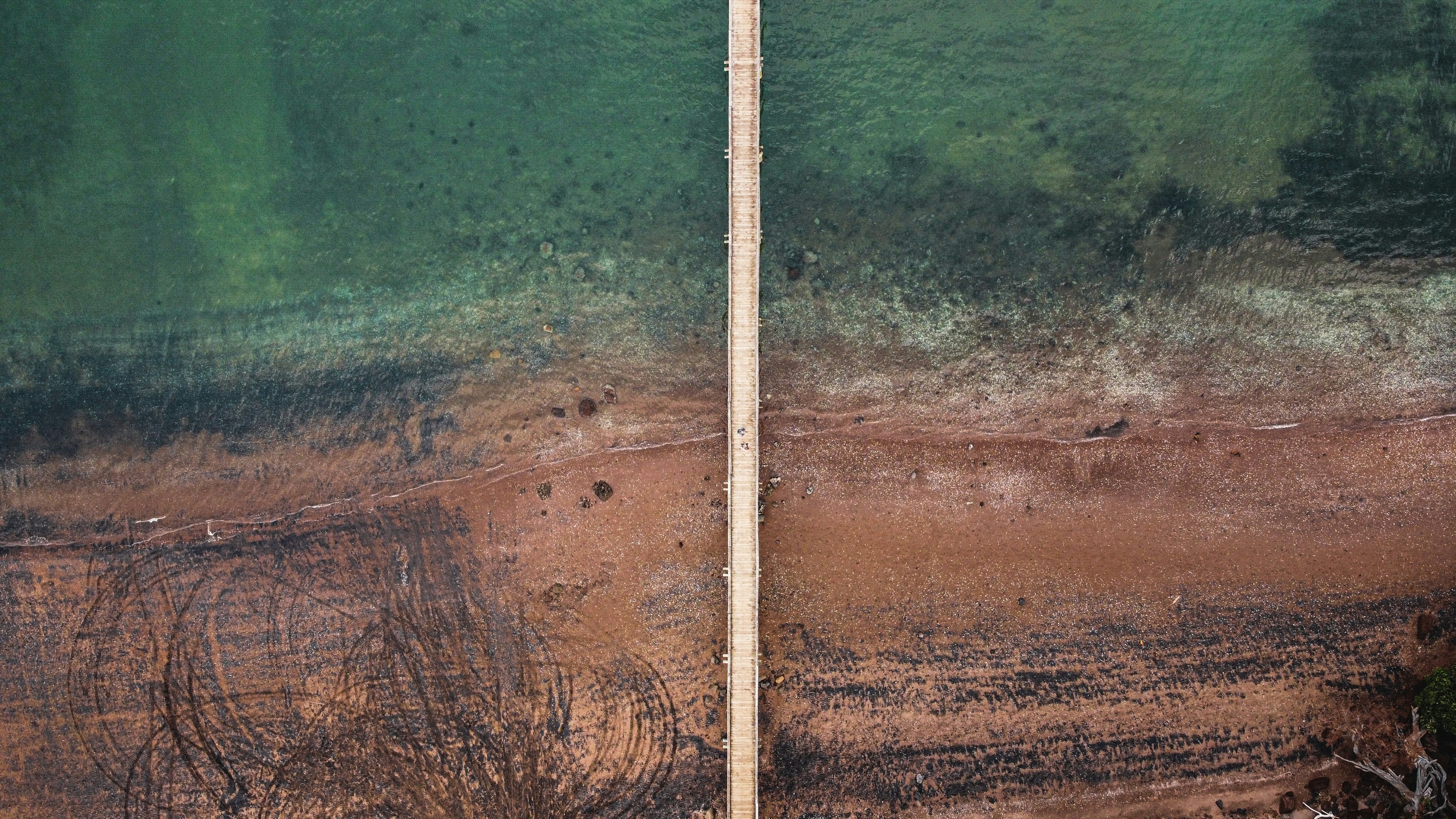 Aerial view of a long wooden bridge stretching across a calm sea, bordered by sandy shores and gentle waves. The contrasting textures of sand and water create a serene coastal scene.