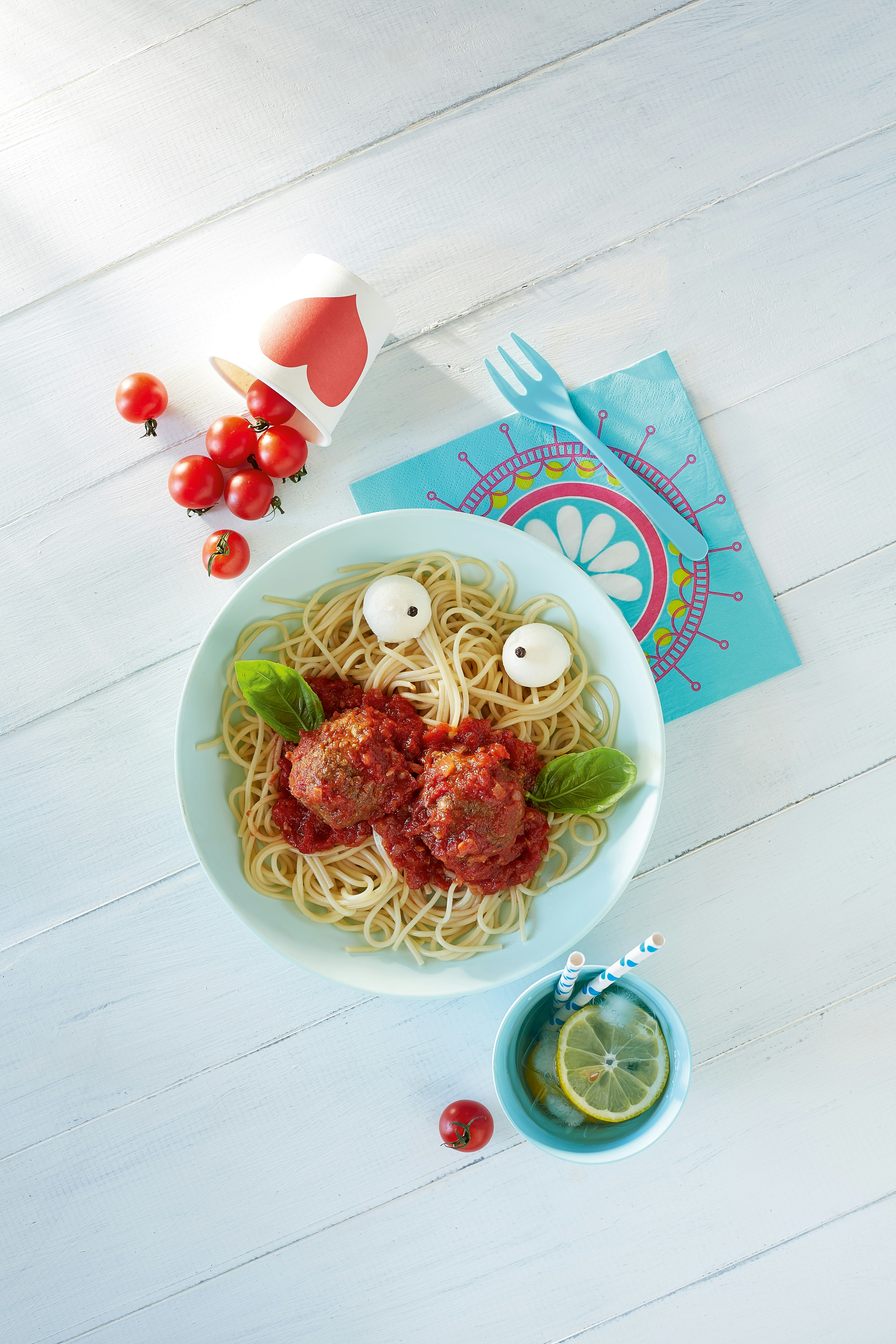Photograph of a bowl of spaghetti with meatballs and two cheese-eye garnishes on a white wooden table, with a blue citrus drink nearby and a decorative napkin. The scene emphasizes whimsy in a homey meal.