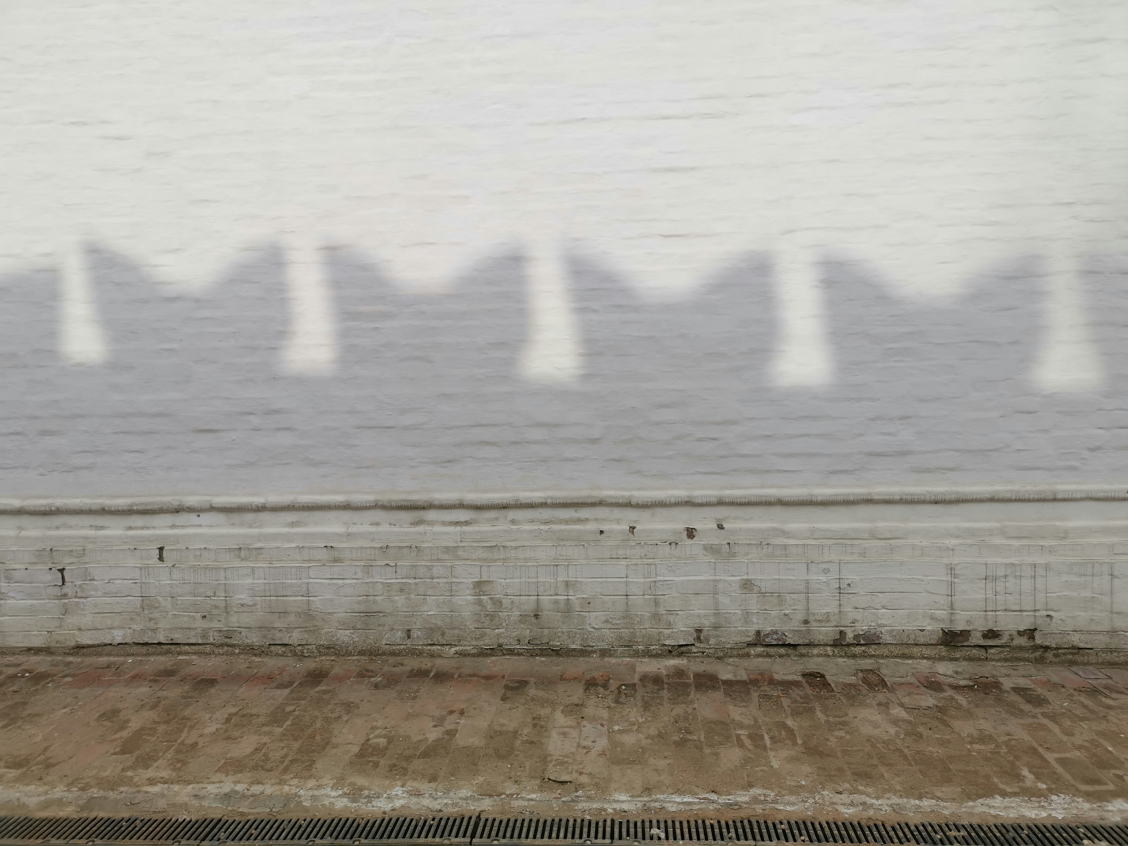 Muted wall and worn wooden base meet a brick-tiled floor in a subdued interior. Repetitive triangular shadows create a geometric pattern across the wall.