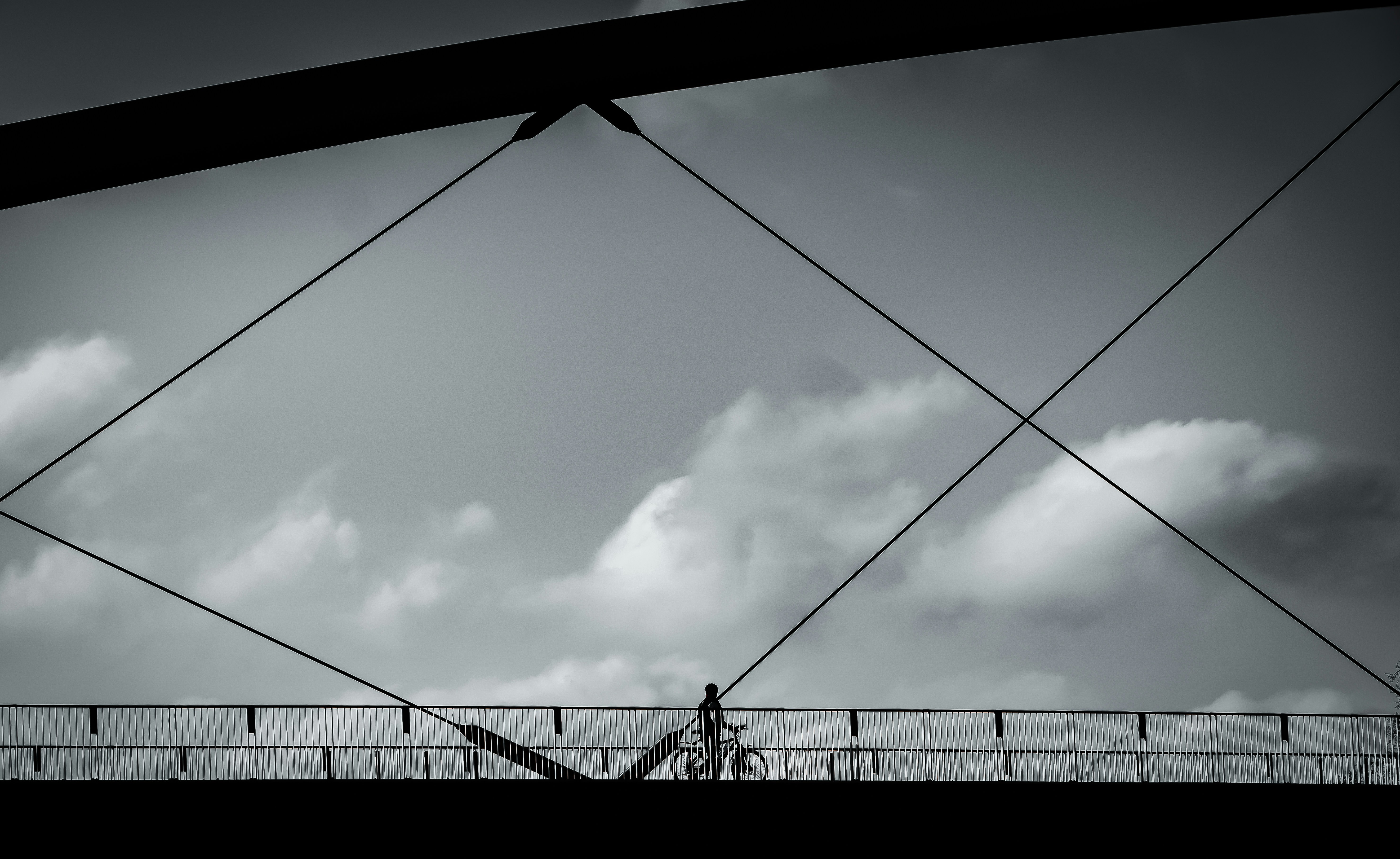 A solitary figure stands on a bridge, framed by the geometric lines of its structure against a dramatic sky. The monochromatic tones enhance the sense of isolation.