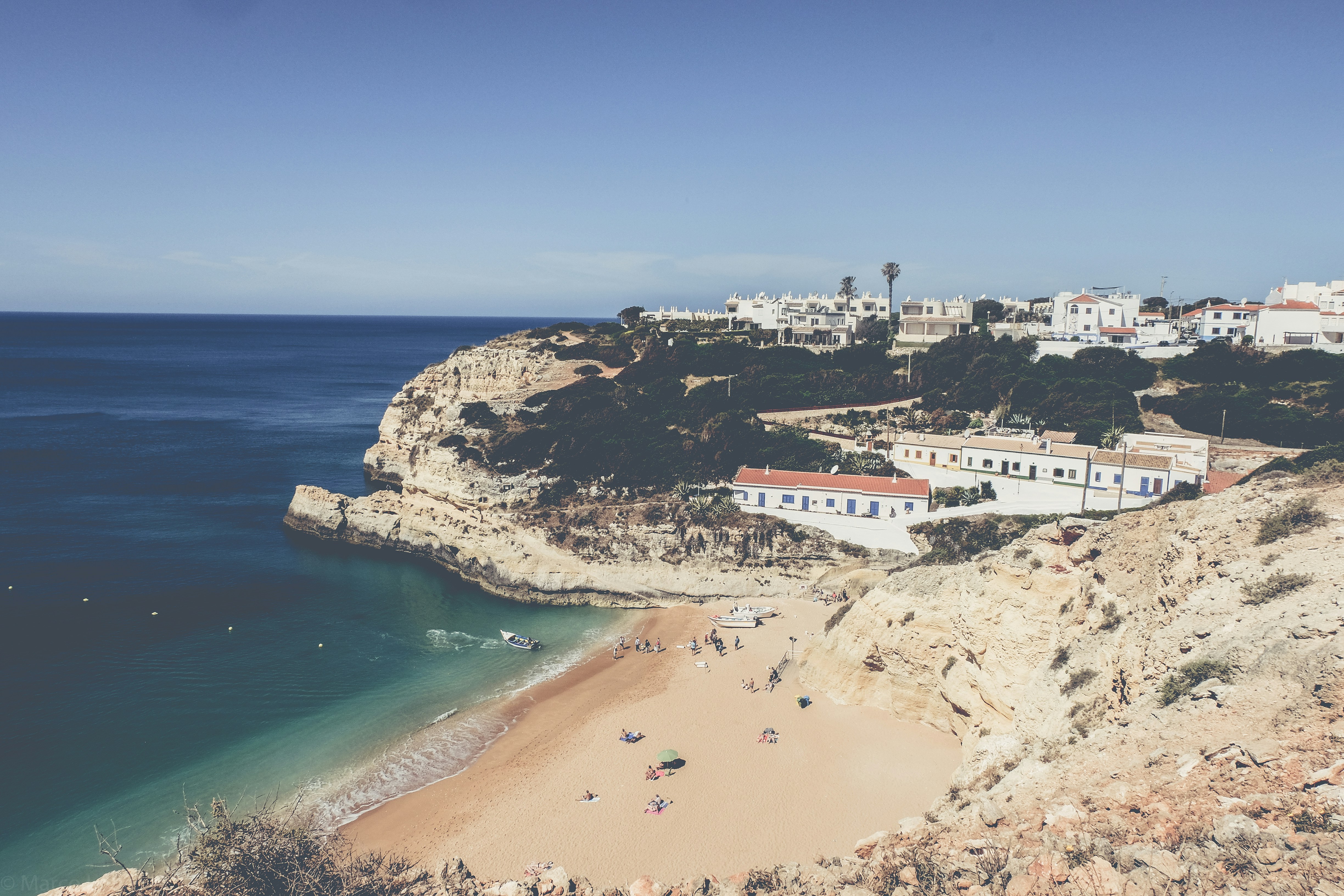 Coastal landscape with sandy beach, rugged cliffs, and white buildings beneath a clear blue sky.