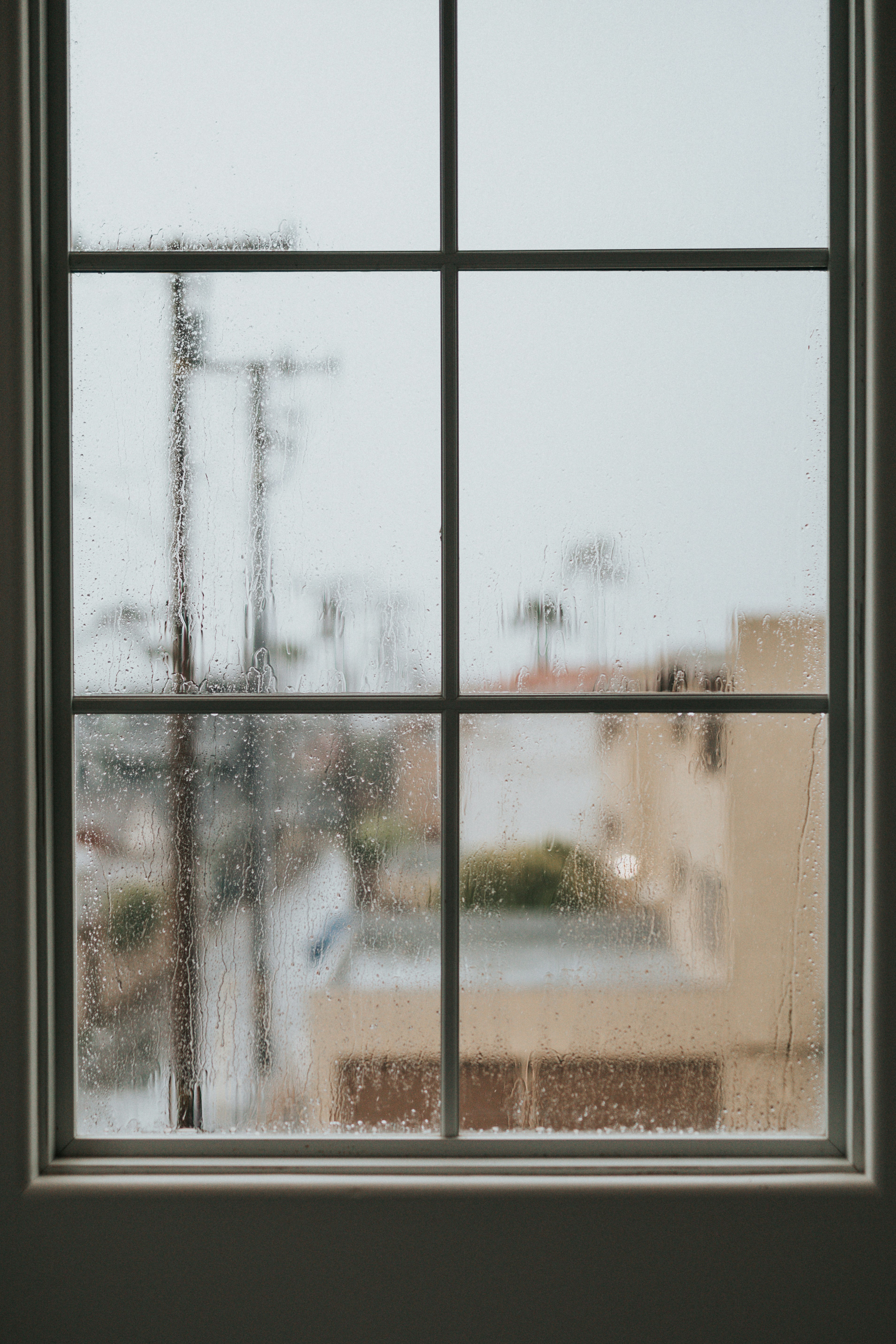 View through a rain-speckled window, capturing blurred outlines of buildings and utility poles outside. The atmosphere conveys a sense of solitude and introspection.