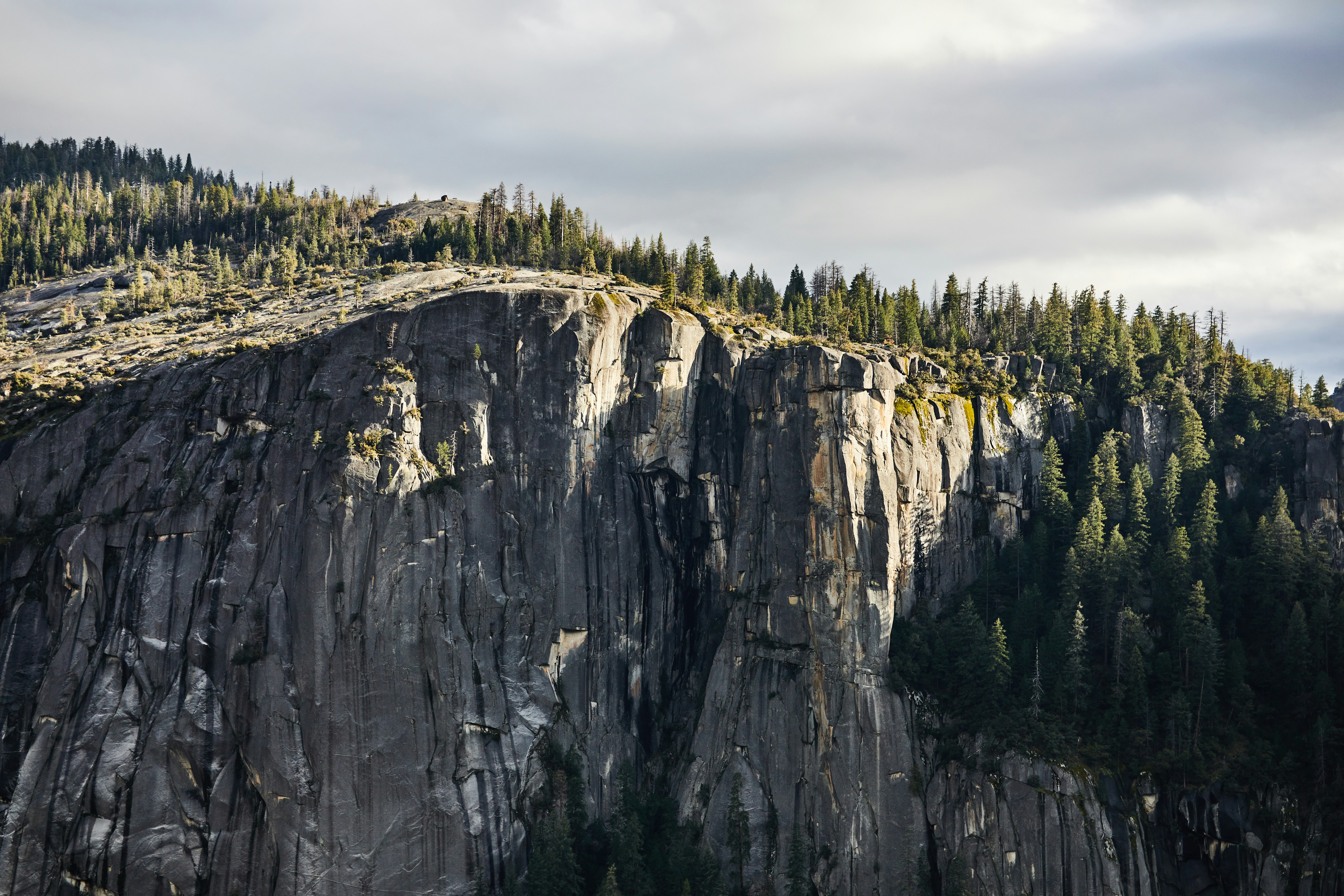 Massive granite cliff rising sharply, adorned with patches of evergreen trees under a cloudy sky.
