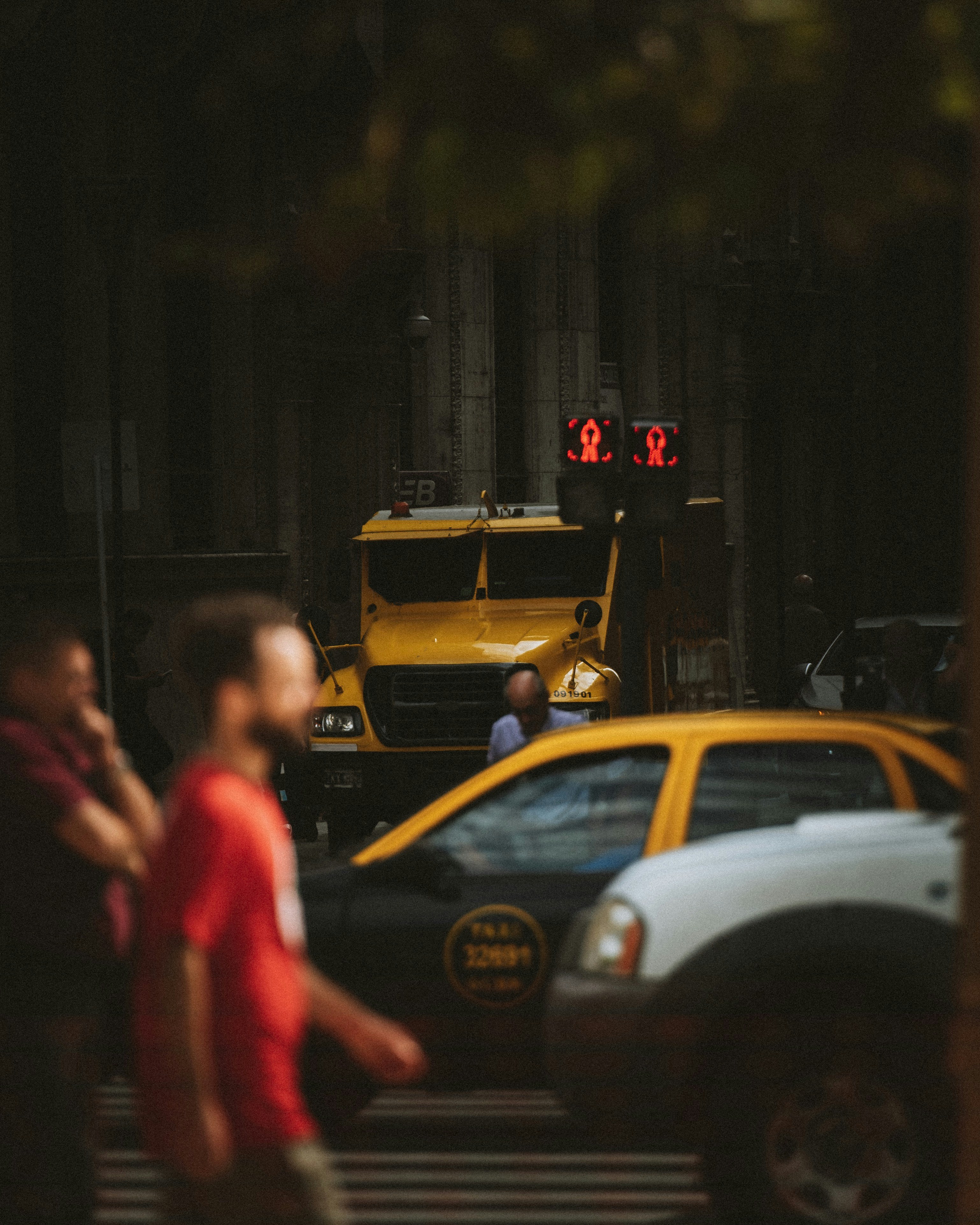 A yellow truck and a taxi are juxtaposed against blurred pedestrians crossing a busy street. Traffic signals and urban architecture create a dynamic city scene.