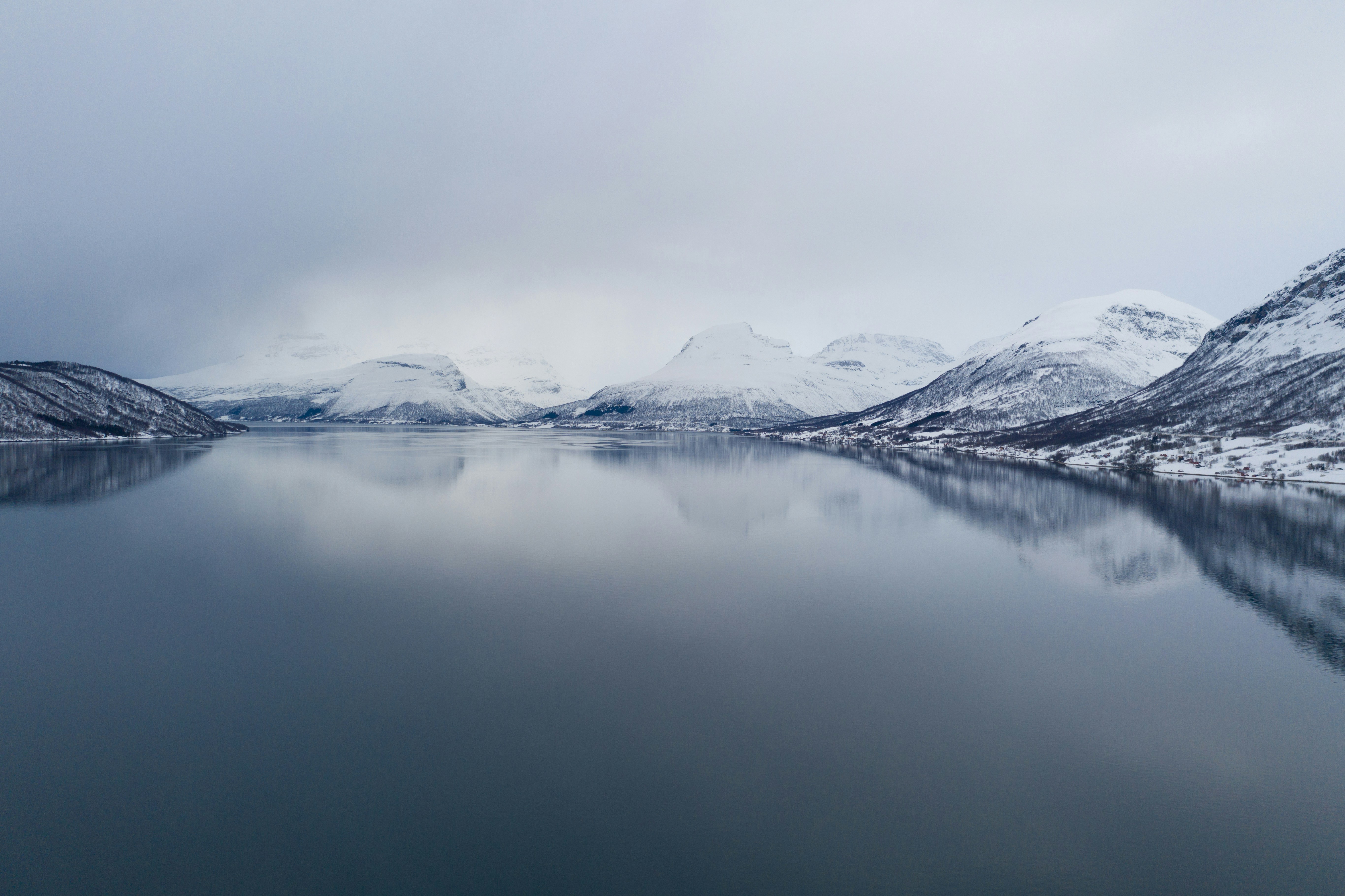 Snow-covered mountains mirrored in a calm fjord under a cloudy sky.