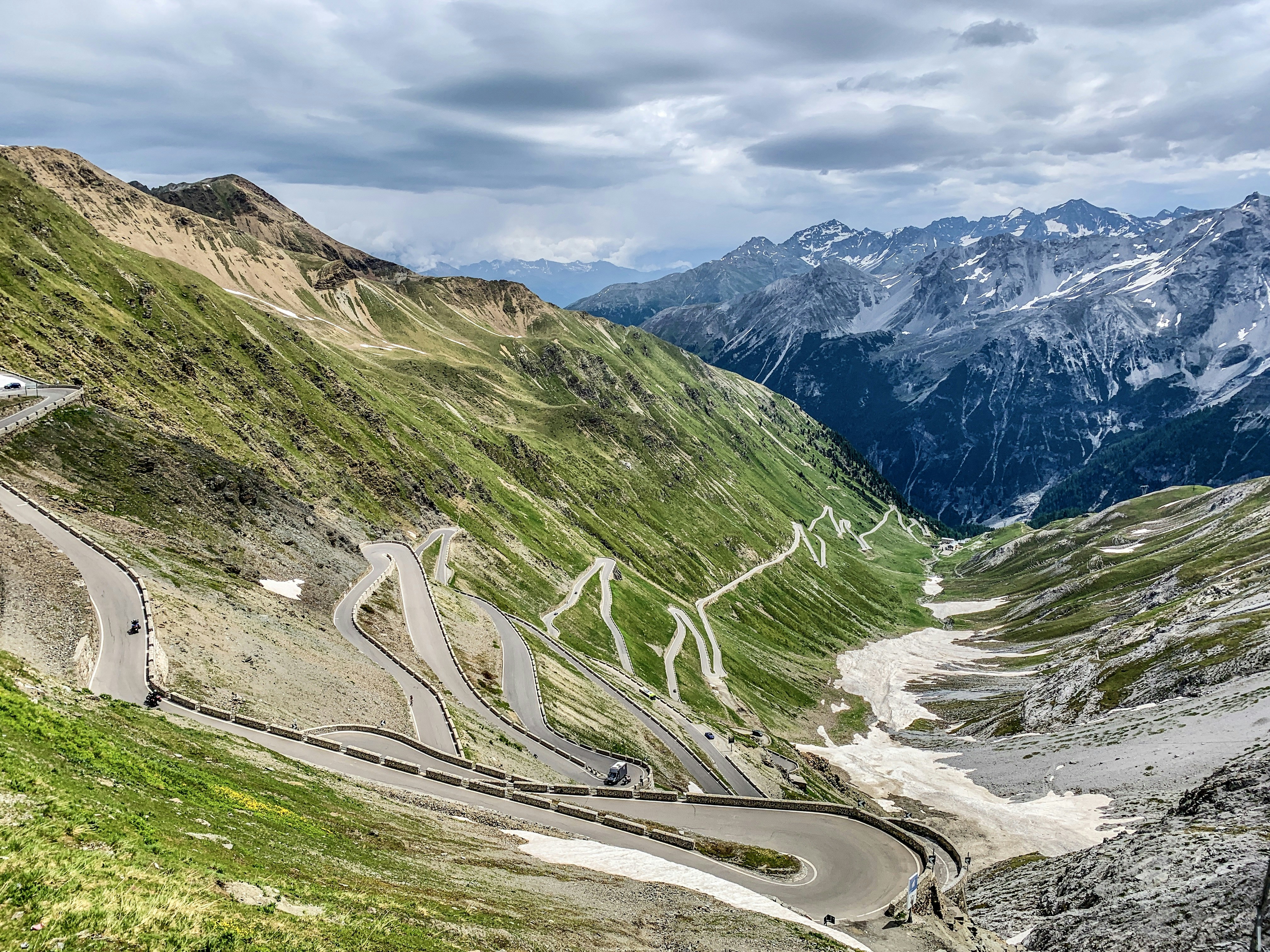 Curving mountain road traverses lush green slopes under a dramatic cloudy sky.