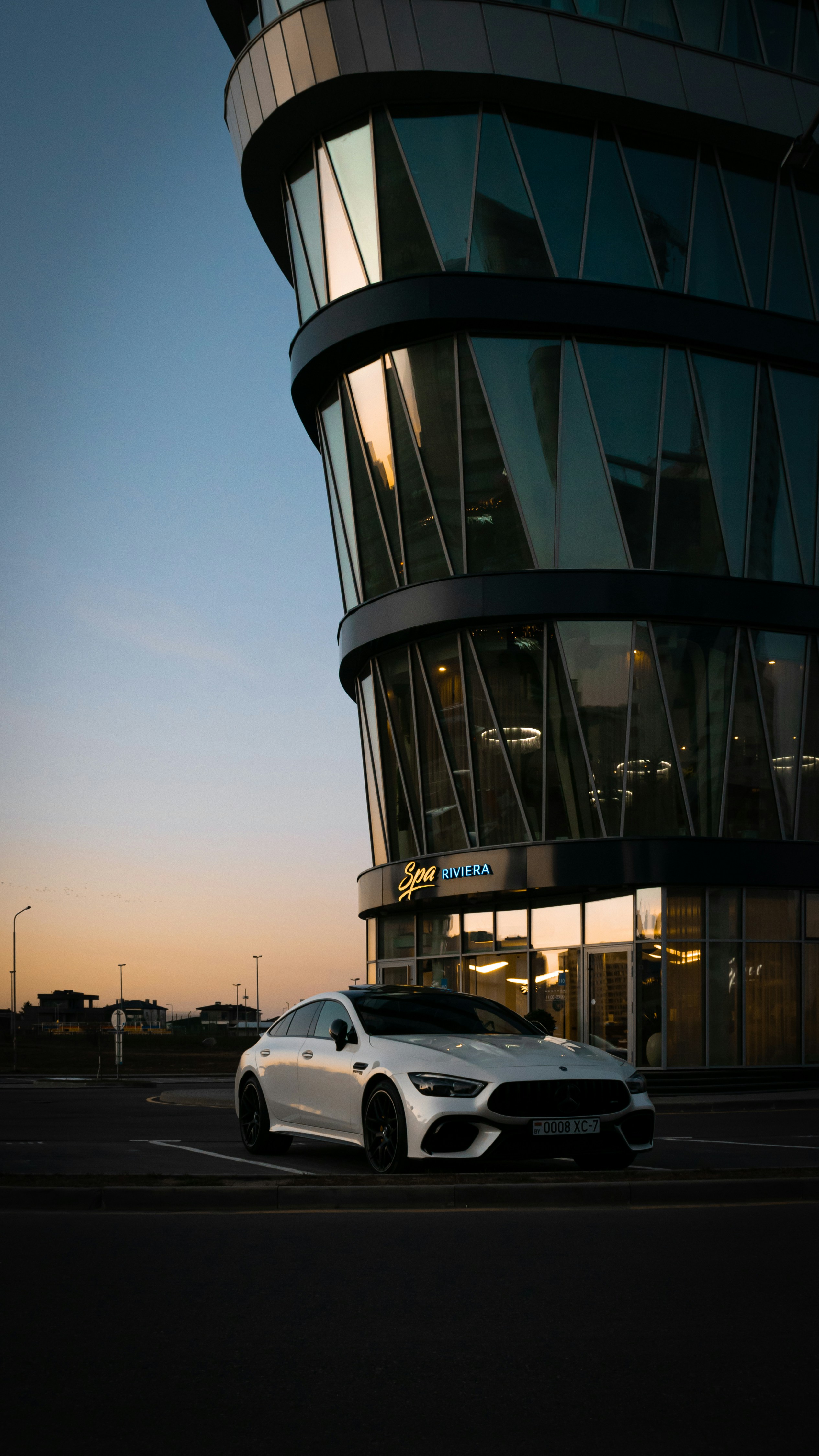 Sleek white sports car parked in front of a contemporary glass building, illuminated by soft twilight. The architectural design reflects the evening sky.