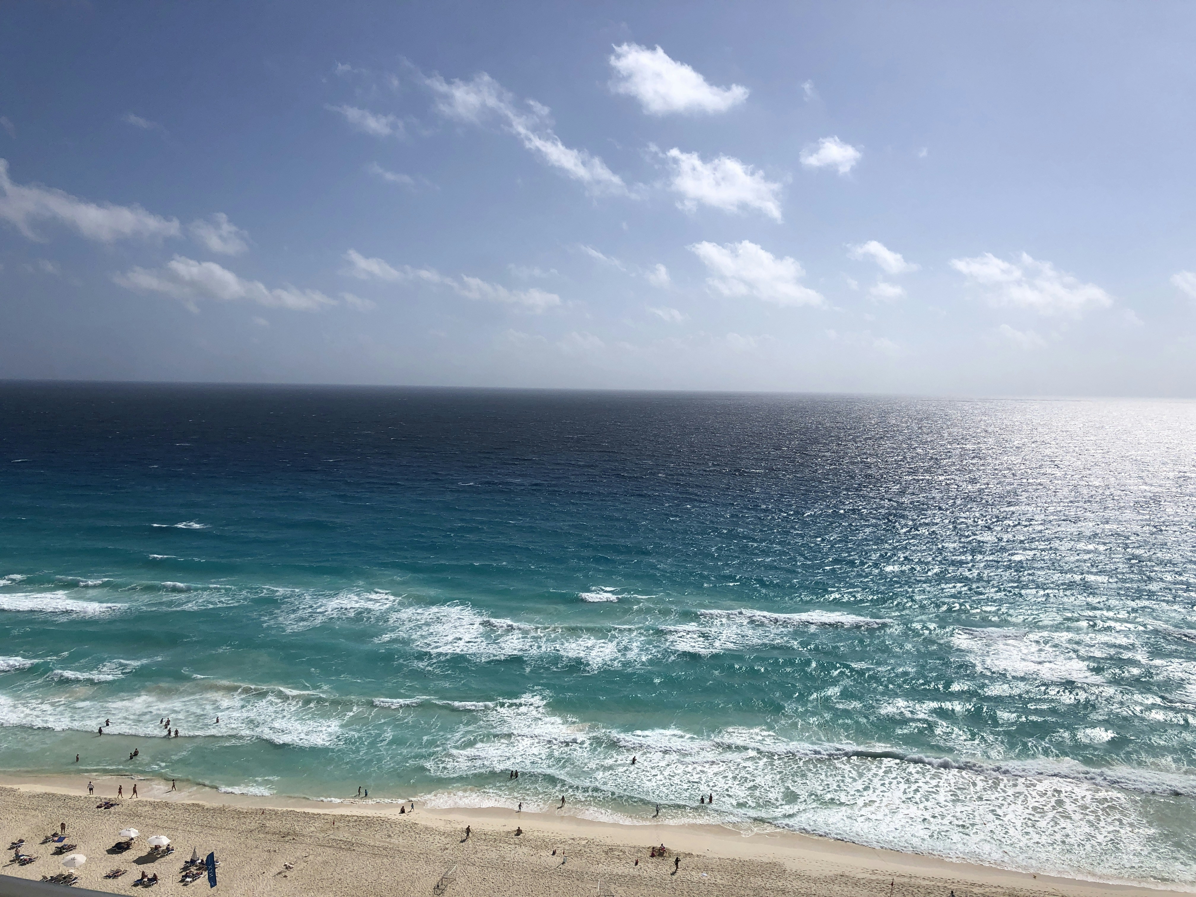 Vast ocean waves gently rolling onto a sandy beach under a bright sky, with scattered beachgoers enjoying the scenery.