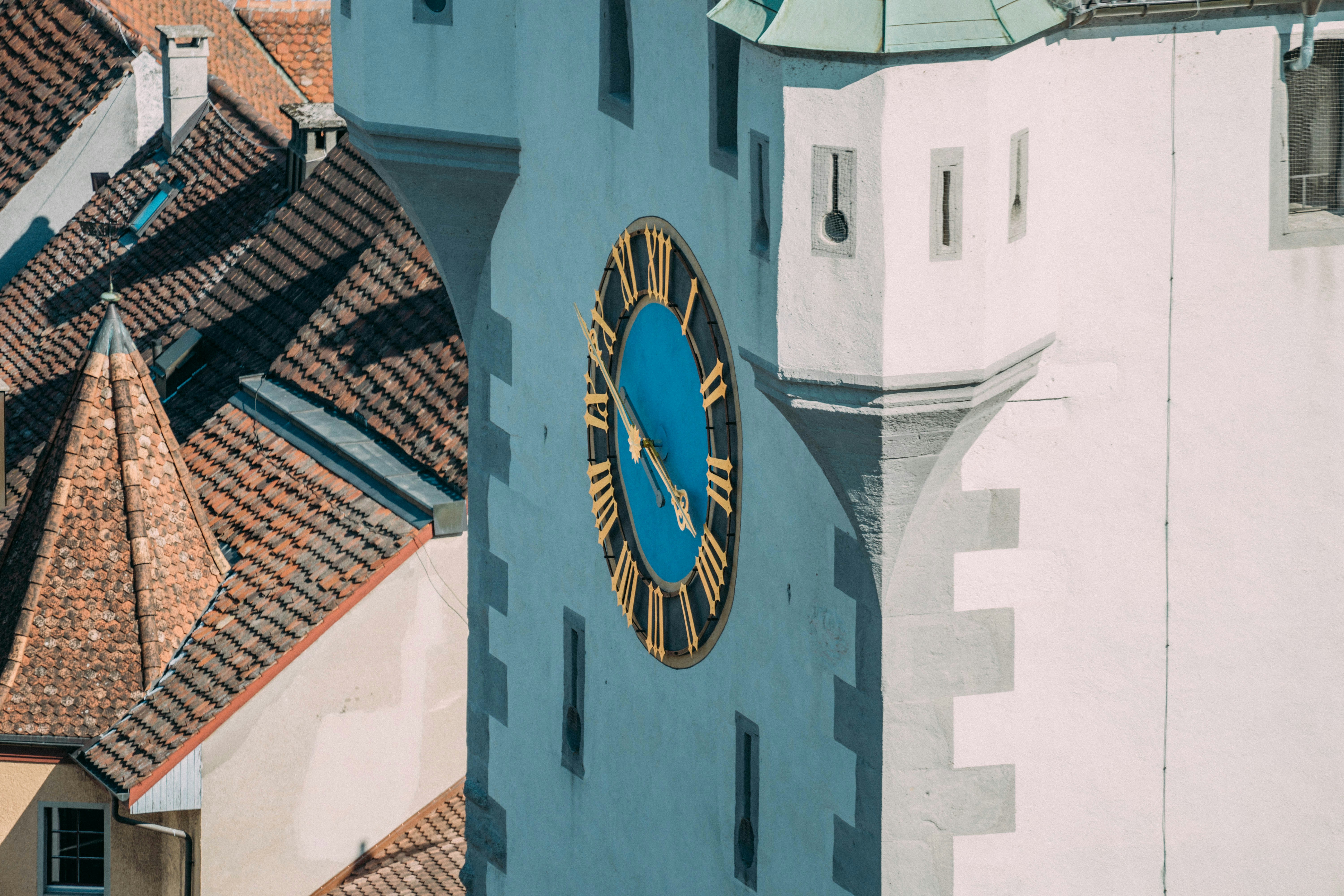Blue and white clock face with Roman numerals on a historic tower beside terracotta rooftops.