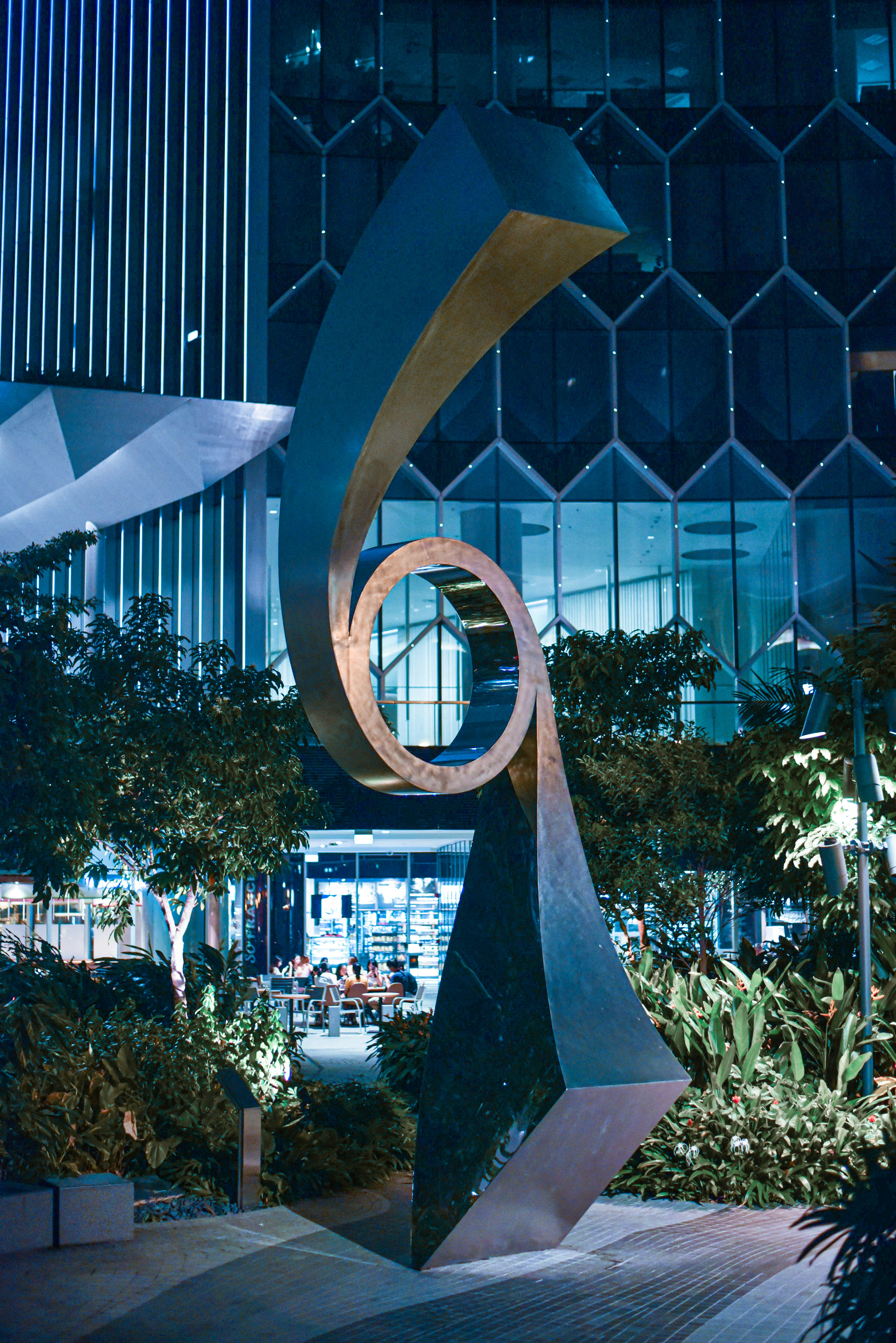 Sculptural representation of a swirling form amidst urban greenery and modern architecture, emphasizing the blend of nature and art in a city landscape.