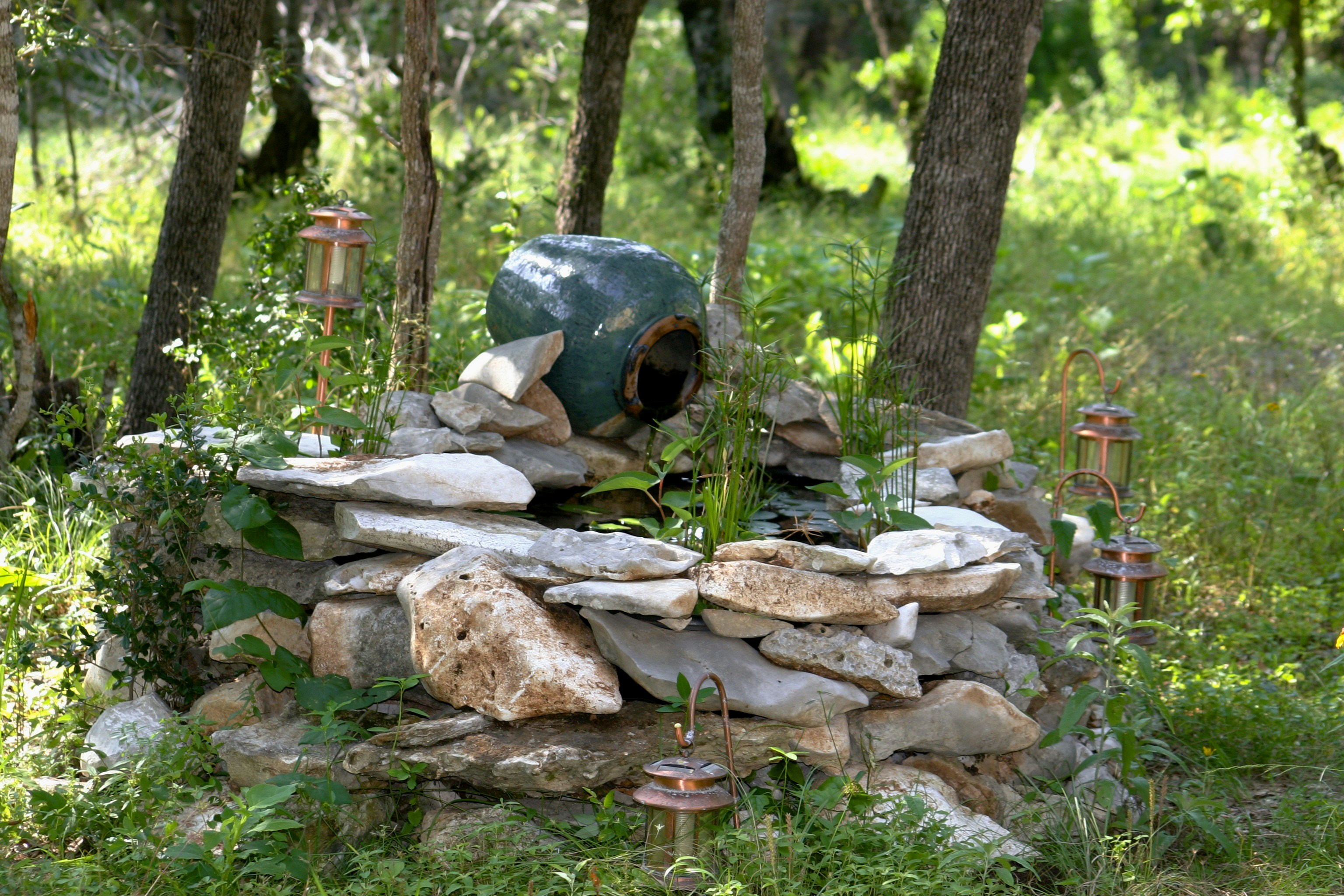 A tranquil stone water feature surrounded by lush greenery, with a ceramic pot and decorative lanterns enhancing the natural setting.