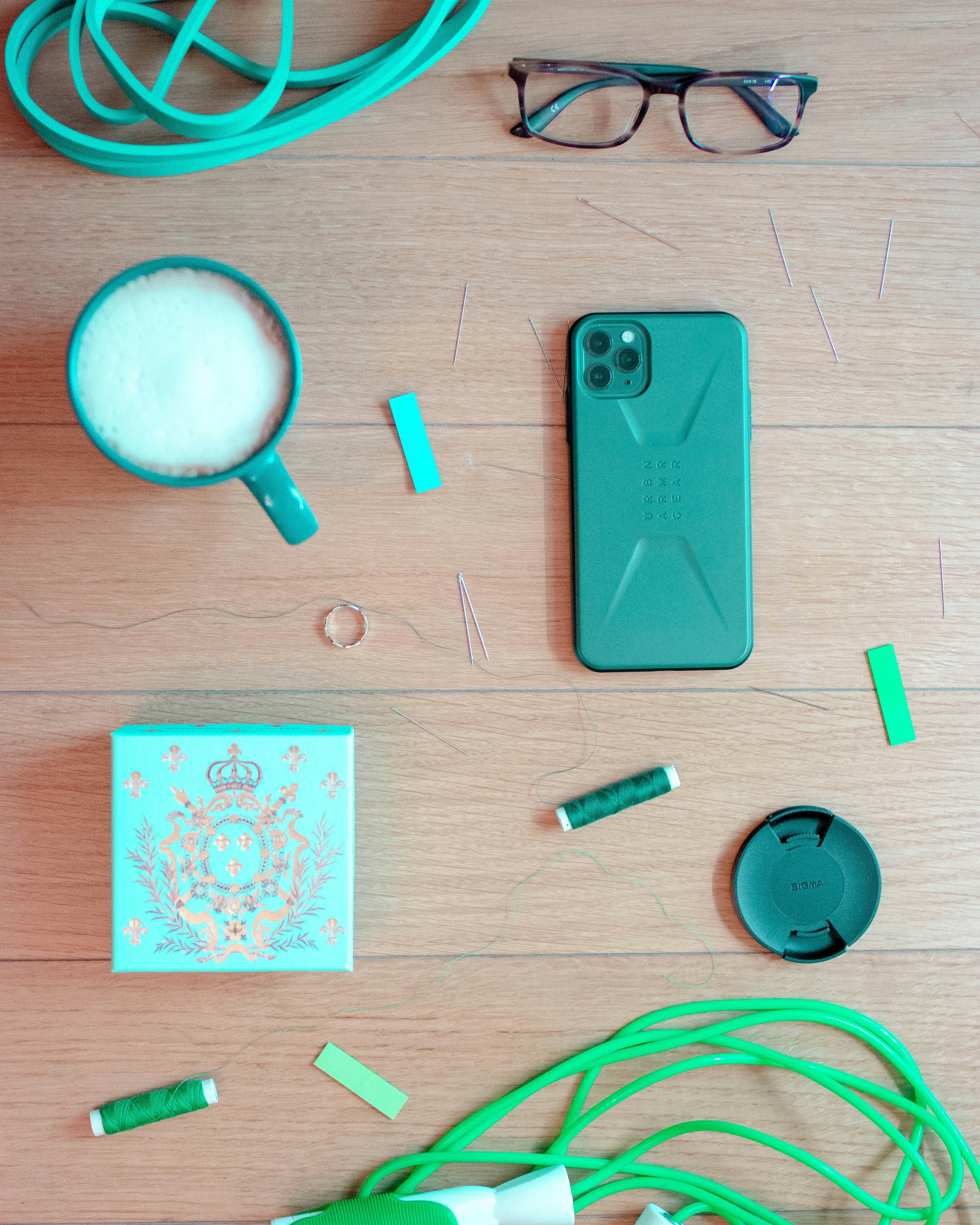 Flat lay arrangement featuring a teal mug, smartphone, sewing supplies, and glasses, all harmoniously placed on a wooden surface.