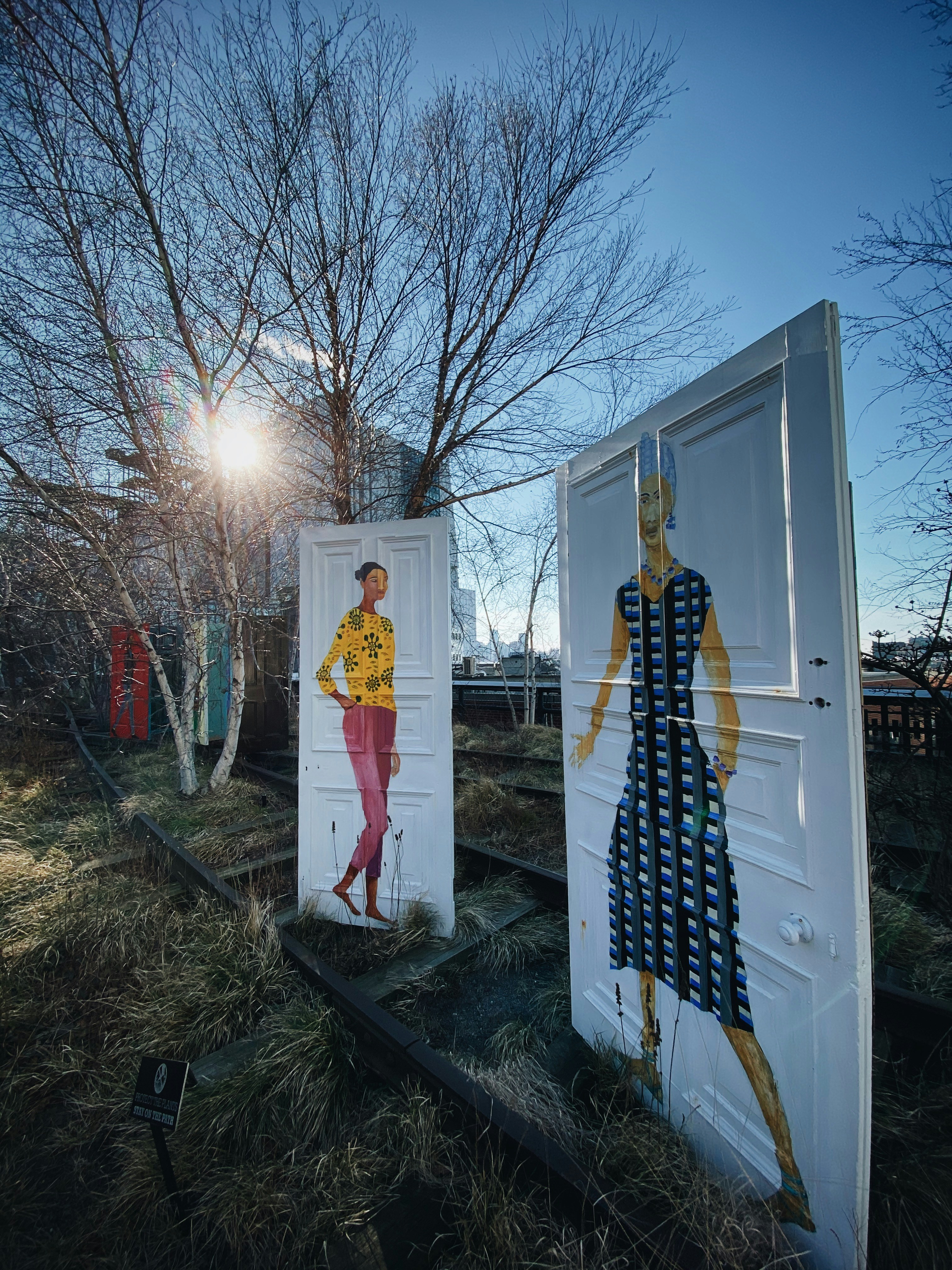Two artistic doors featuring vibrant illustrations of women stand amidst a grassy landscape, with sunlight filtering through nearby trees.