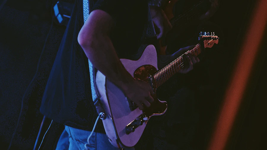 Musician playing an electric guitar on a dimly lit stage with blue and purple lighting. The shot frames the torso, hands, and guitar in a moody live-performance scene.