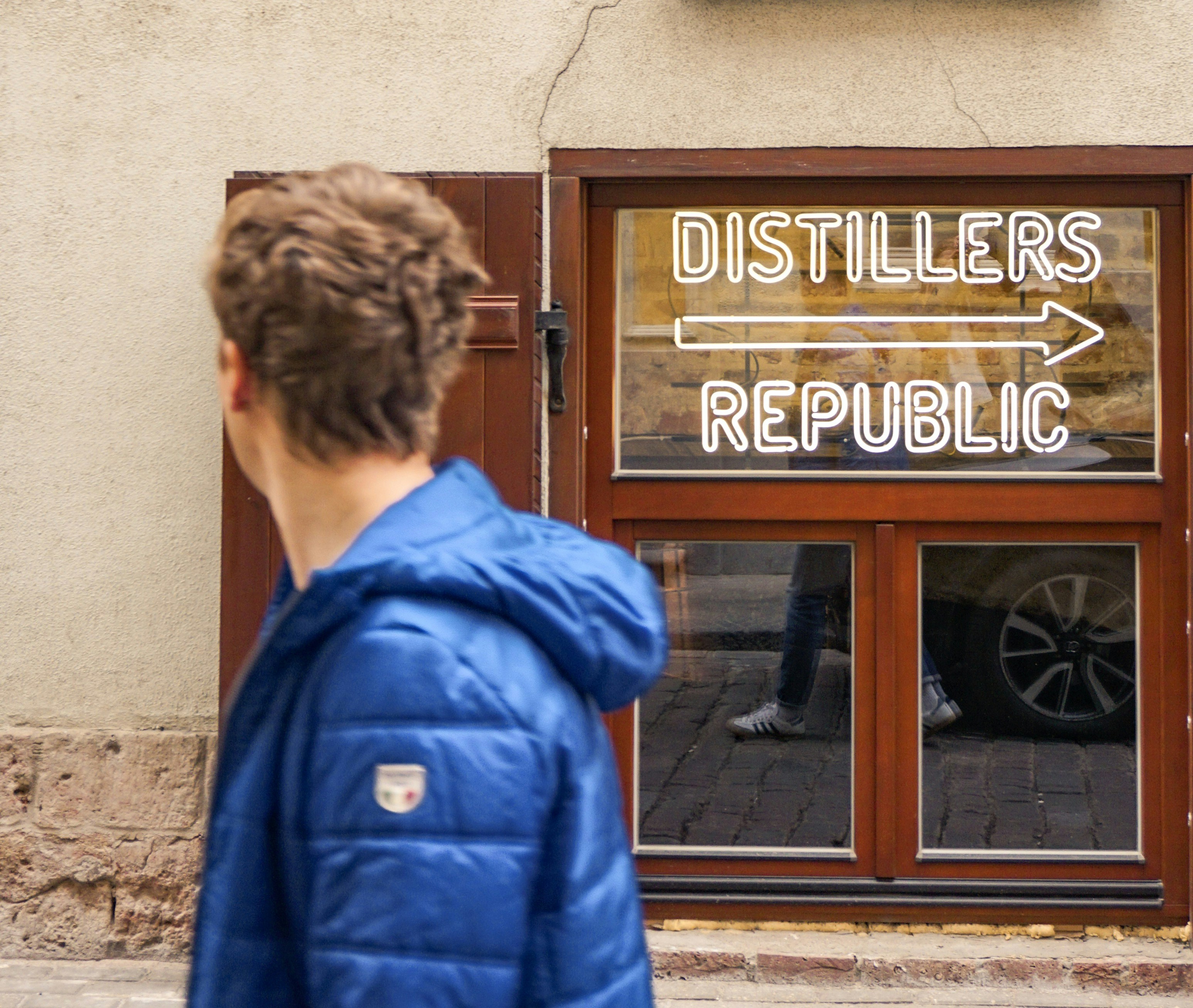 Person in blue jacket glancing toward a sign reading 'Distillers Republic' on a wooden door.