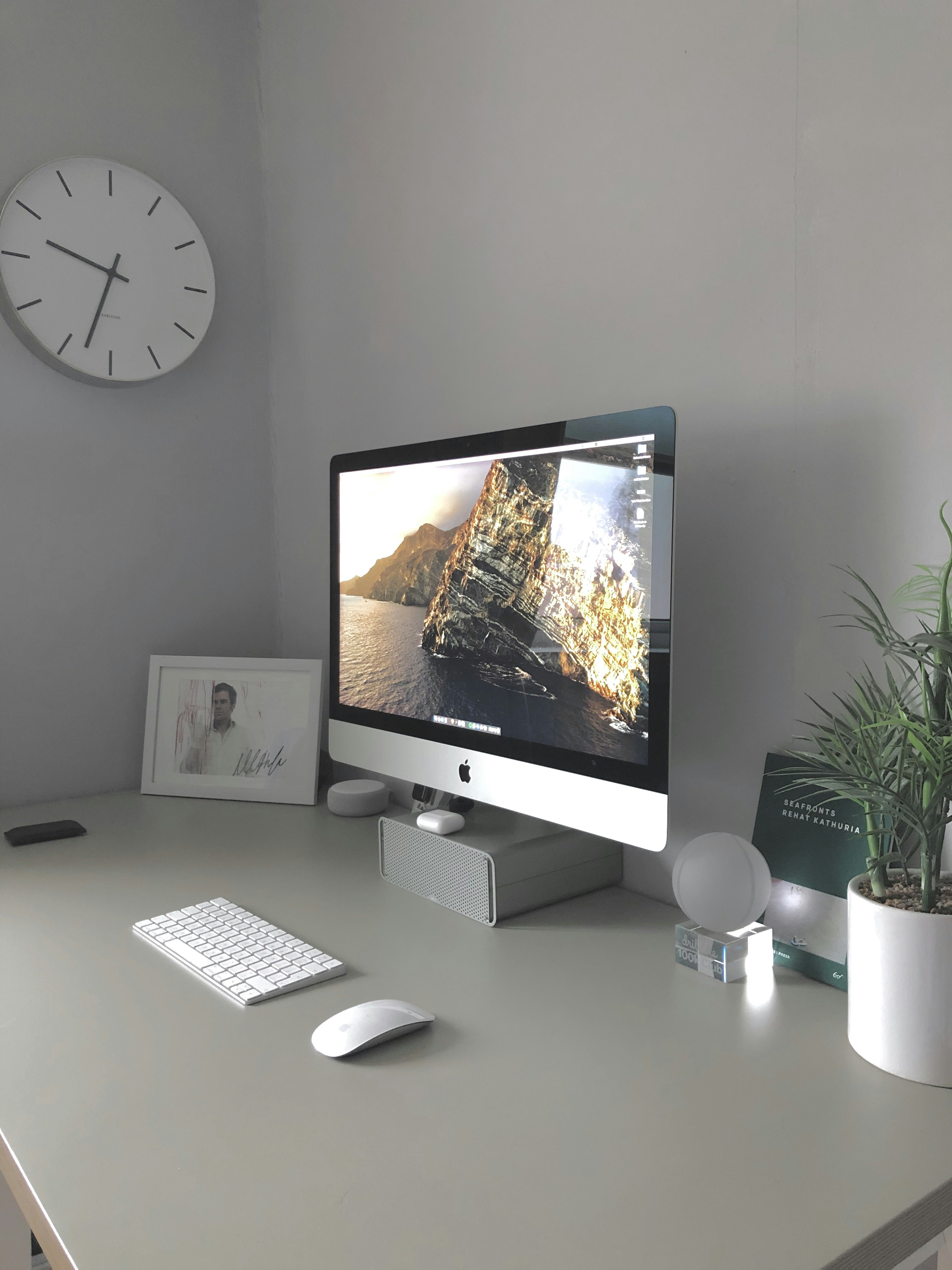 Sleek iMac displaying a scenic image on a minimalist desk with a clock, keyboard, and decorative plant.
