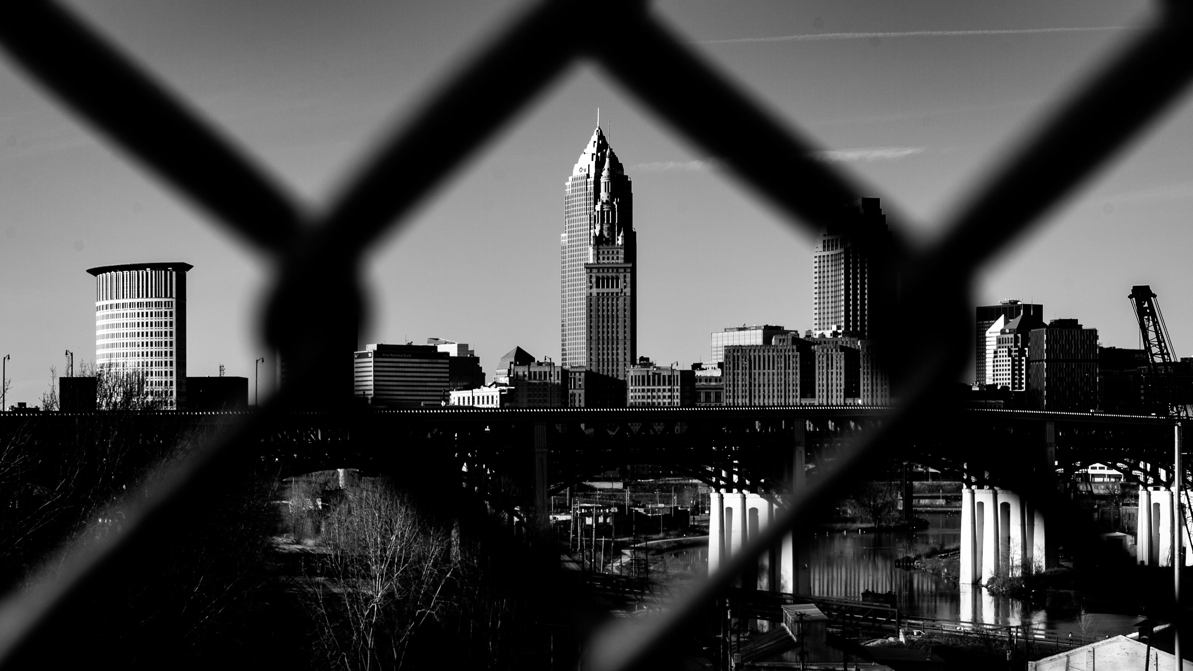 Cleveland's skyline framed by a chain-link fence, highlighting the contrast between urban architecture and industrial elements. Black and white tones emphasize the city's structural lines.