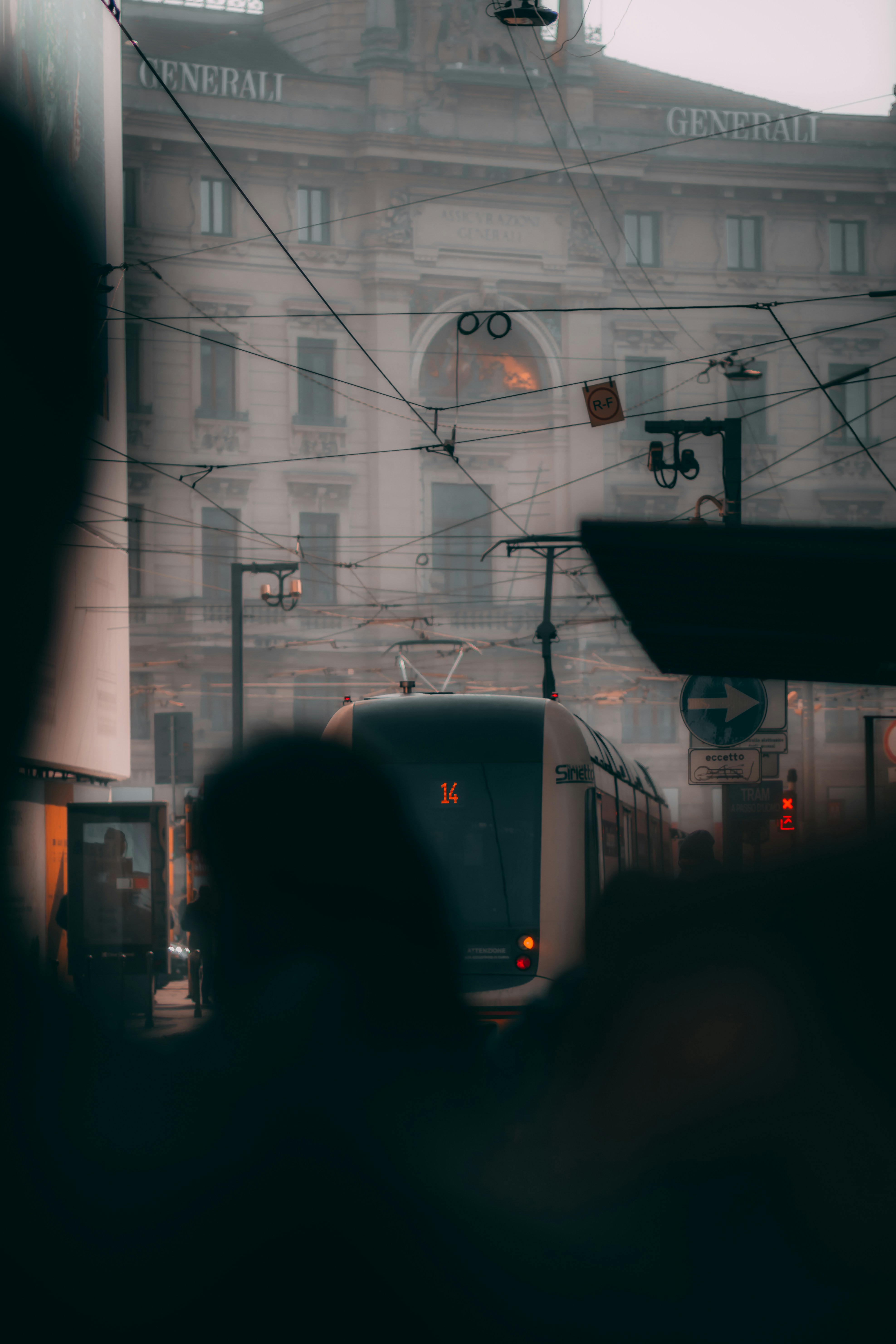 A tram navigates through a foggy urban landscape, with blurred figures of pedestrians creating a sense of movement. Street signs and overhead wires add to the city's intricate web.