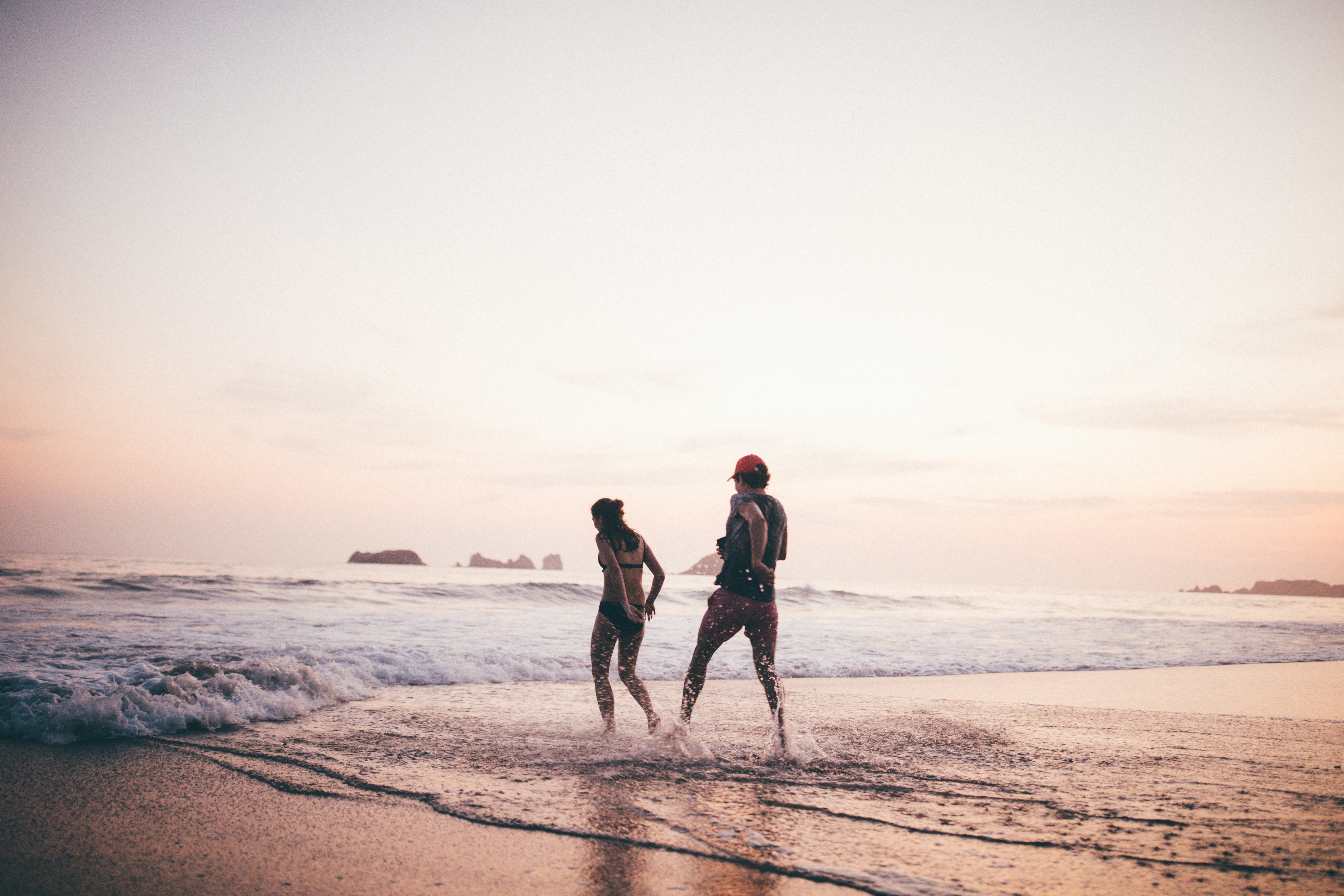 Two figures joyfully running along the shoreline as waves crash against their legs during sunset.