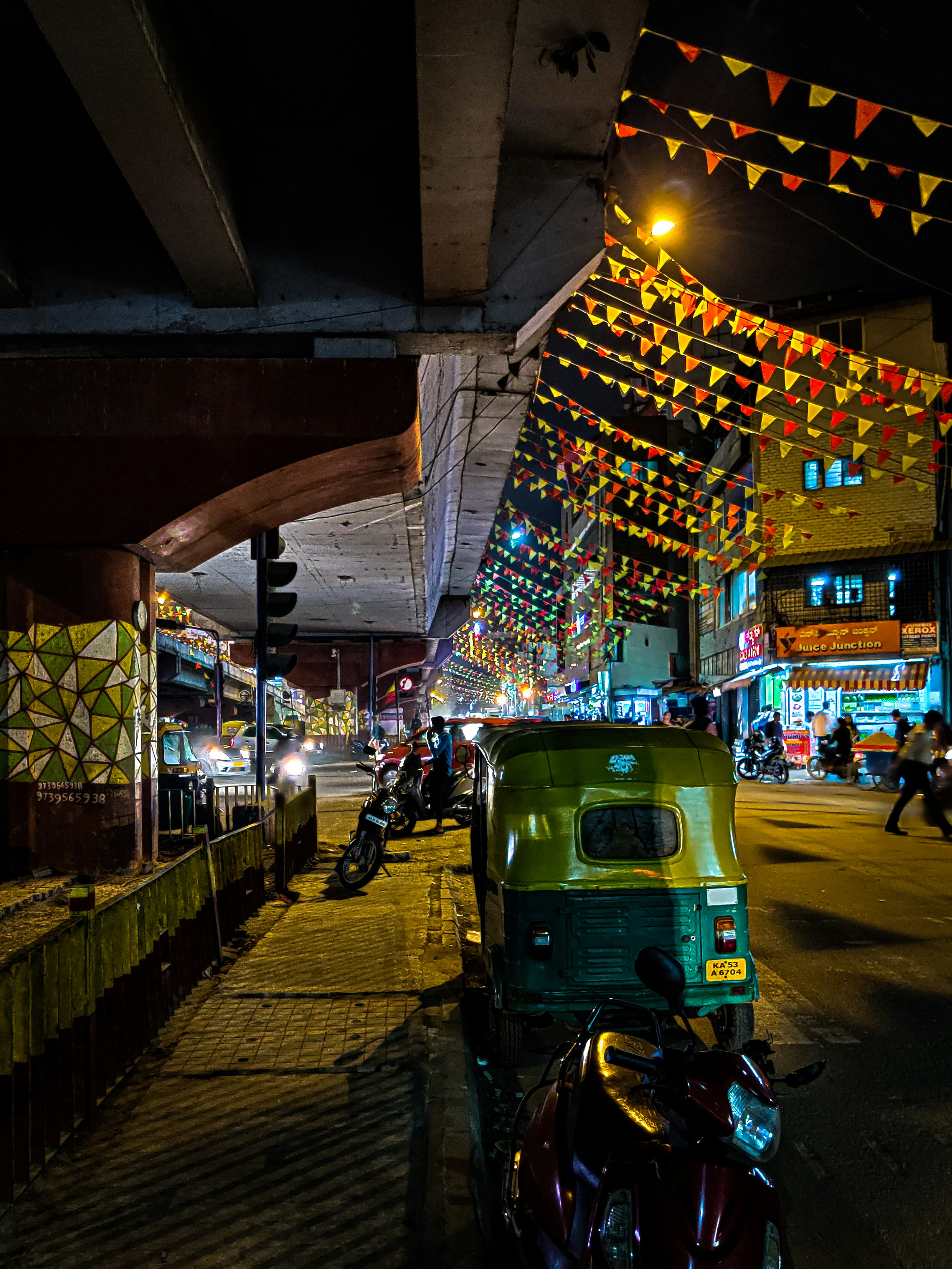 Colorful flags drape across a bustling street scene under an overpass, illuminated by city lights. Auto-rickshaws and motorcycles add to the lively atmosphere.