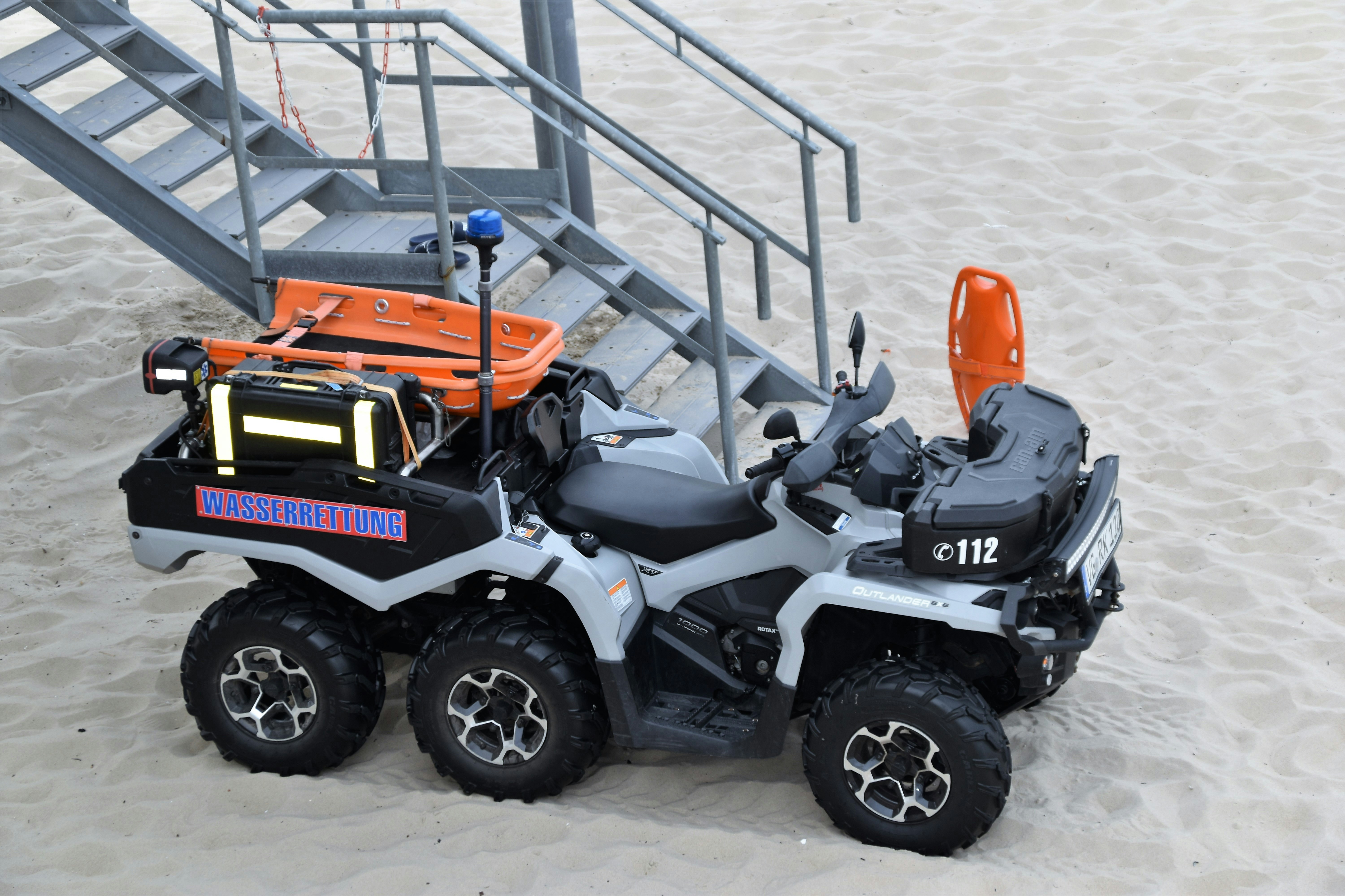 A lifeguard ATV equipped for beach emergencies rests on sandy terrain near a set of stairs. The vehicle features essential rescue gear and is ready for action.