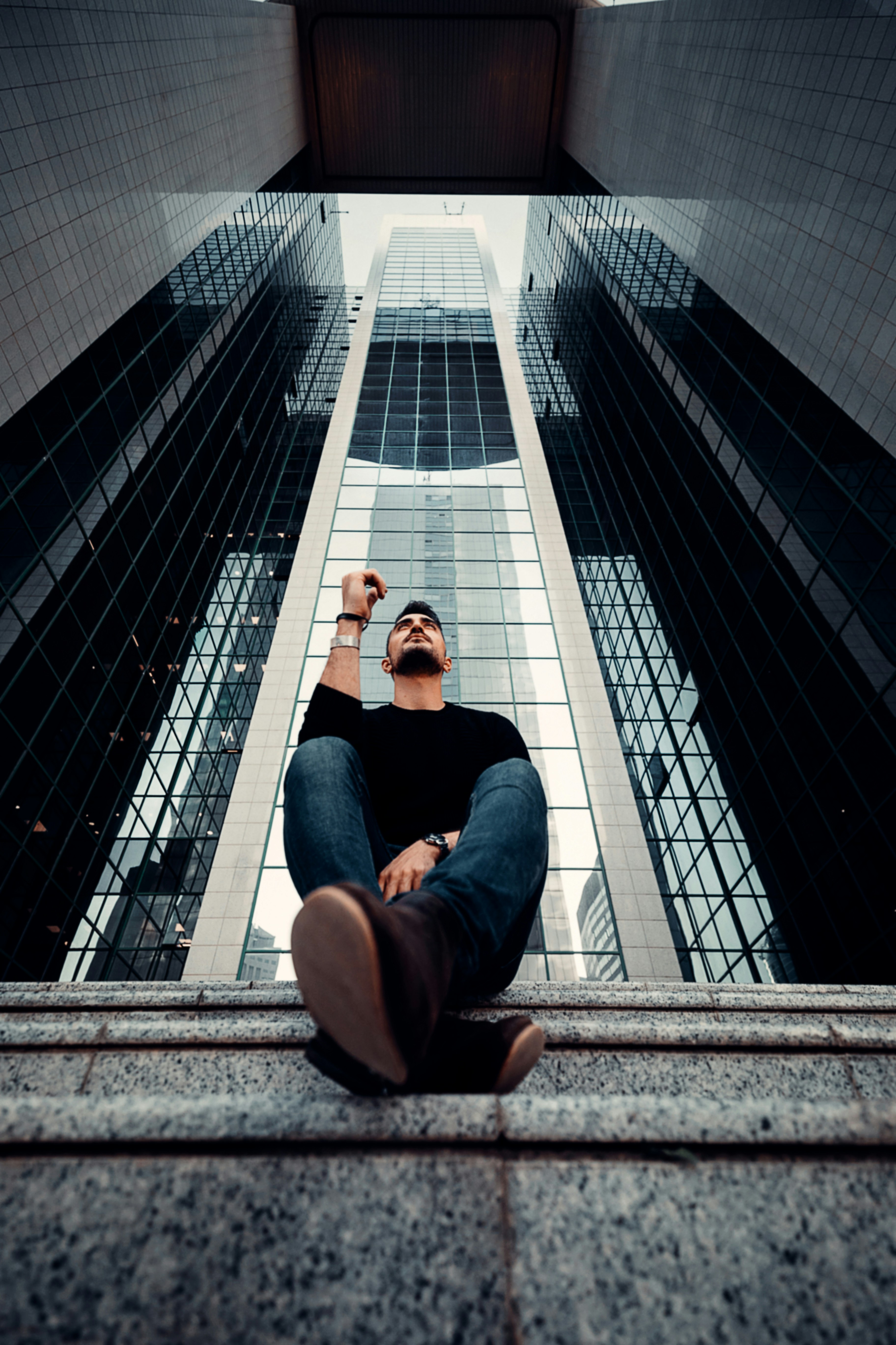 A man sits on stone steps, gazing upward at towering glass buildings, reflecting the urban landscape. The scene captures the contrast between human presence and architectural grandeur.