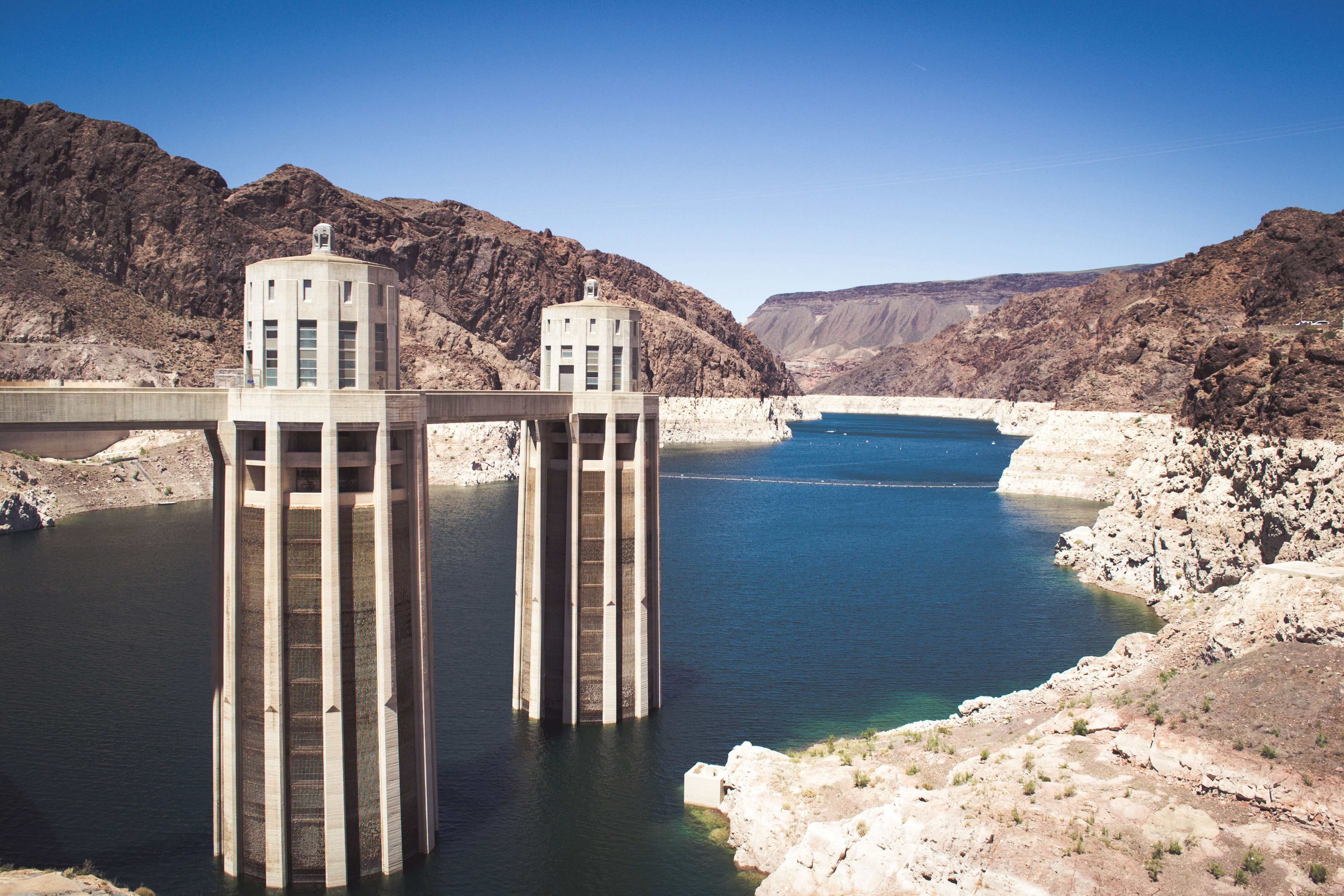 Concrete towers stand sentinel over a vast reservoir, framed by rugged mountains under a clear blue sky.