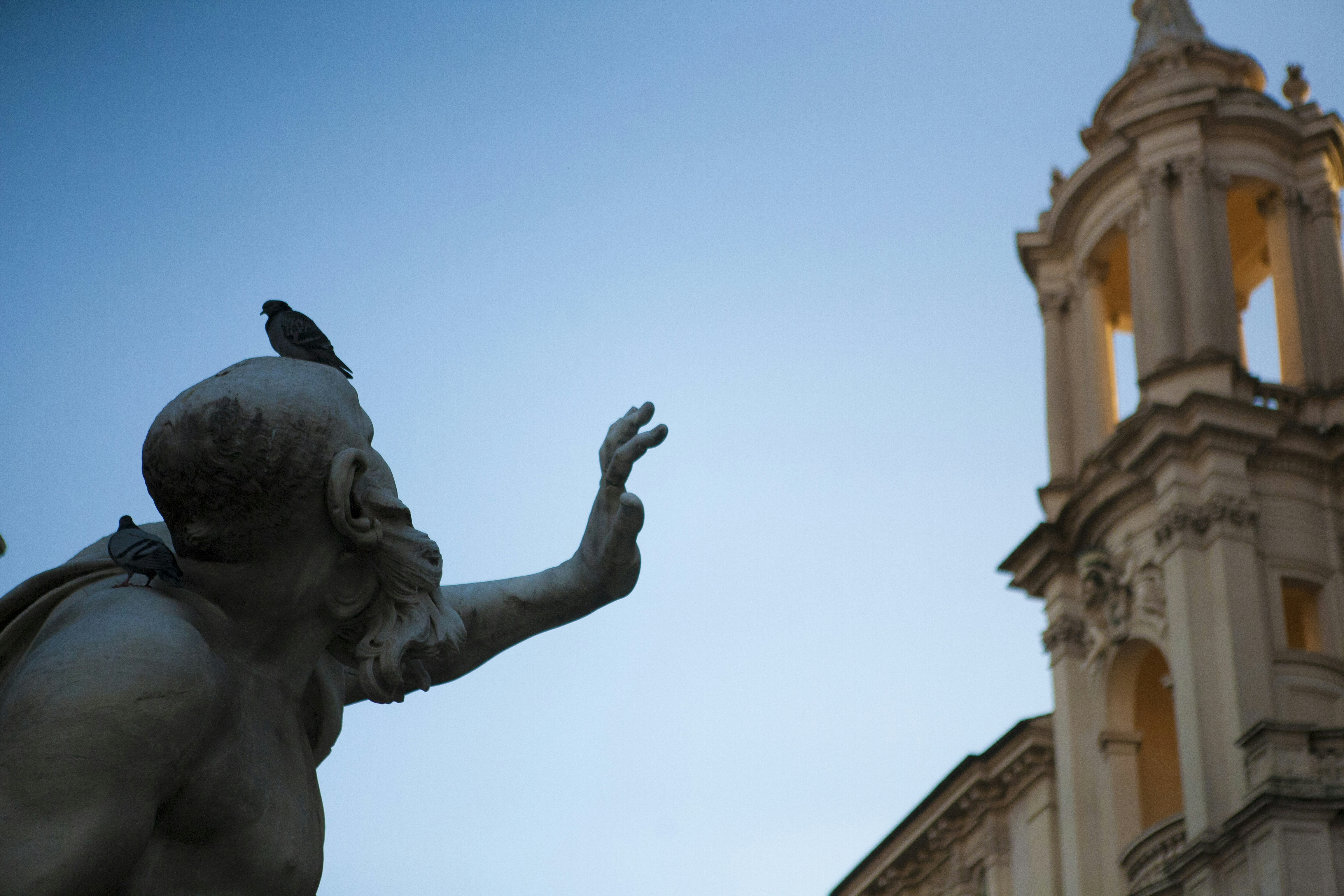 A stone statue of a man reaching out with a bird perched on his head, framed against a clear sky and an ornate building in the background.