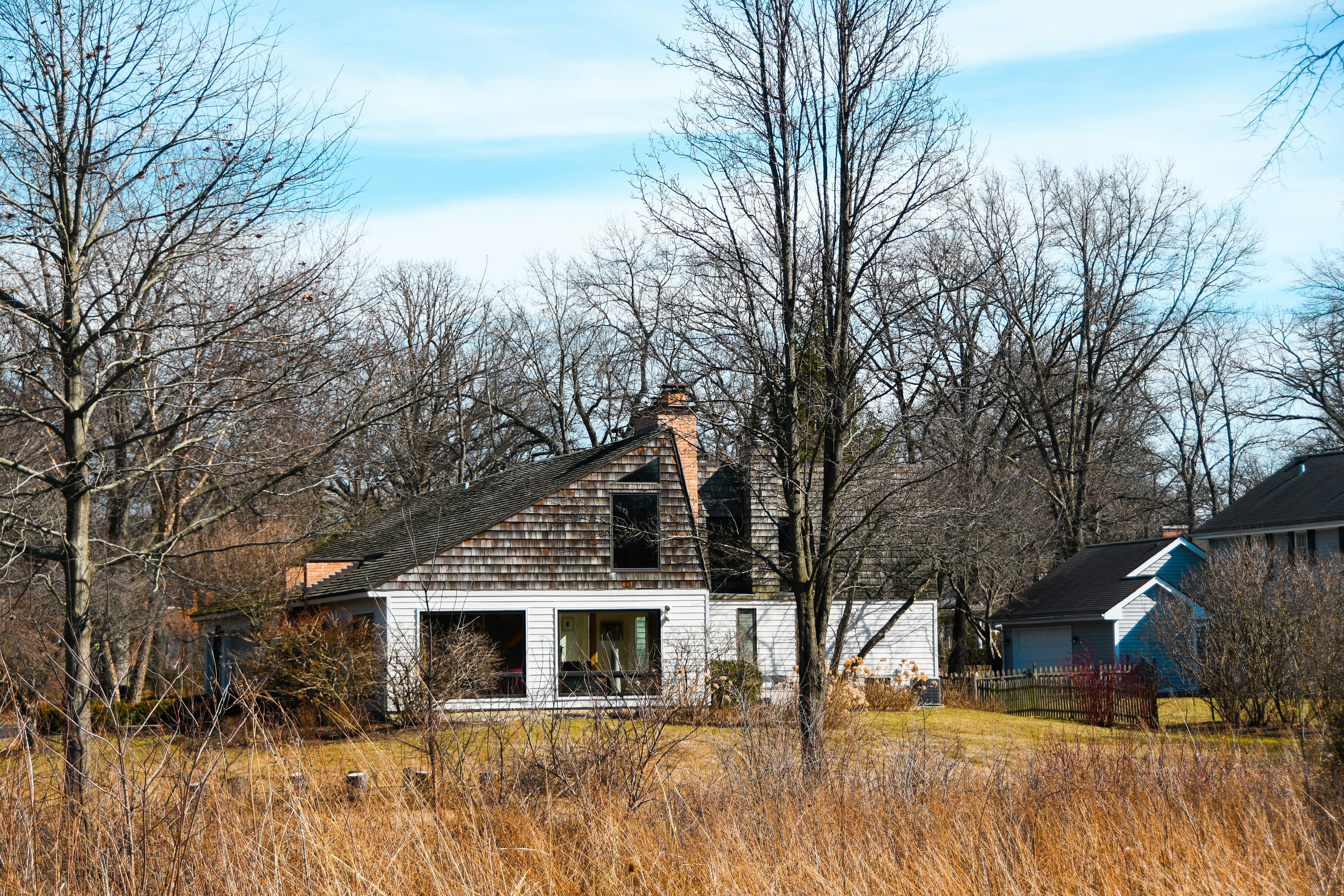 A weathered house with a sloped roof and large windows, surrounded by bare trees and tall grasses, evokes a sense of nostalgia in a tranquil setting.