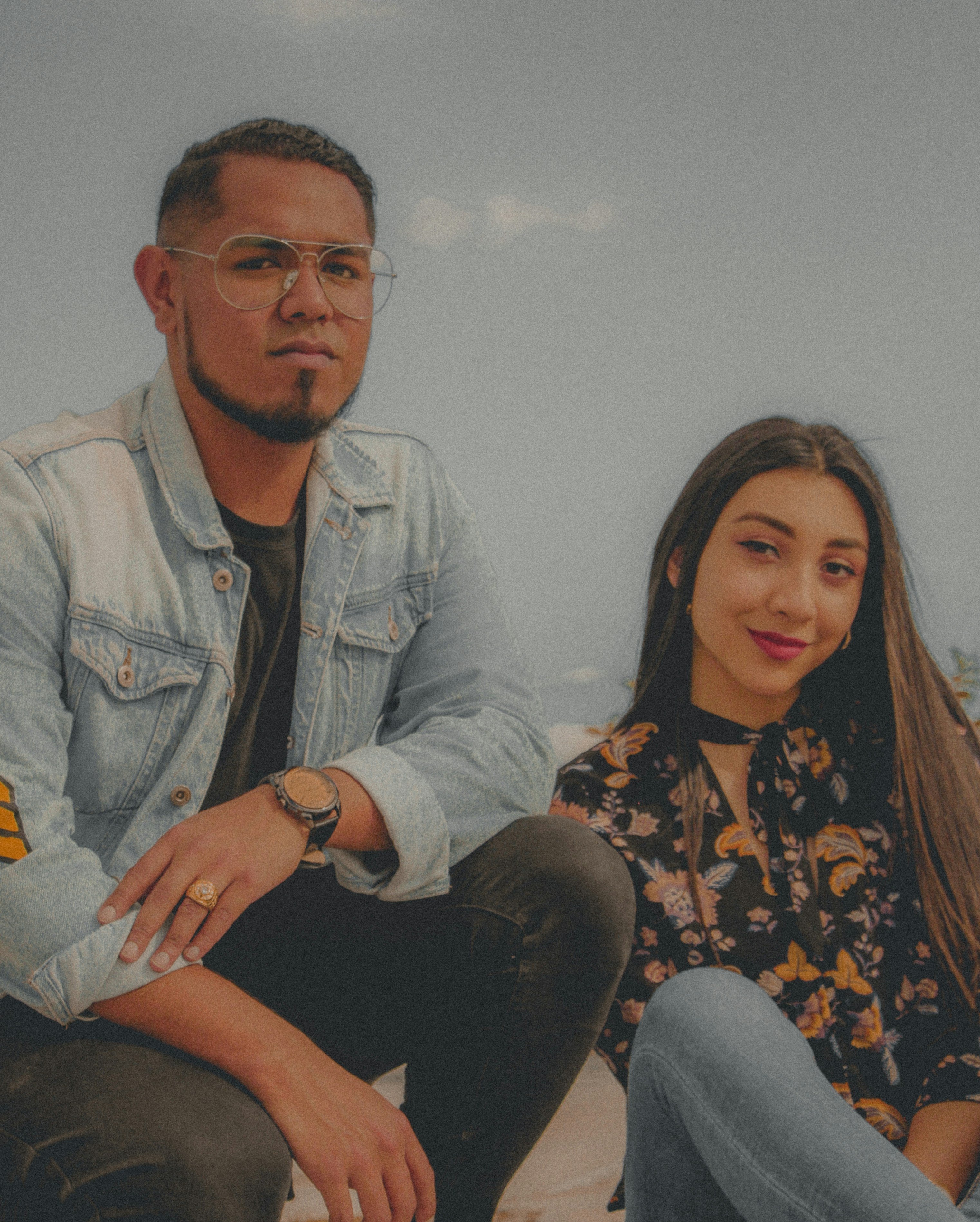 Two stylish individuals pose together against a soft sky backdrop, showcasing their unique fashion and confident expressions.