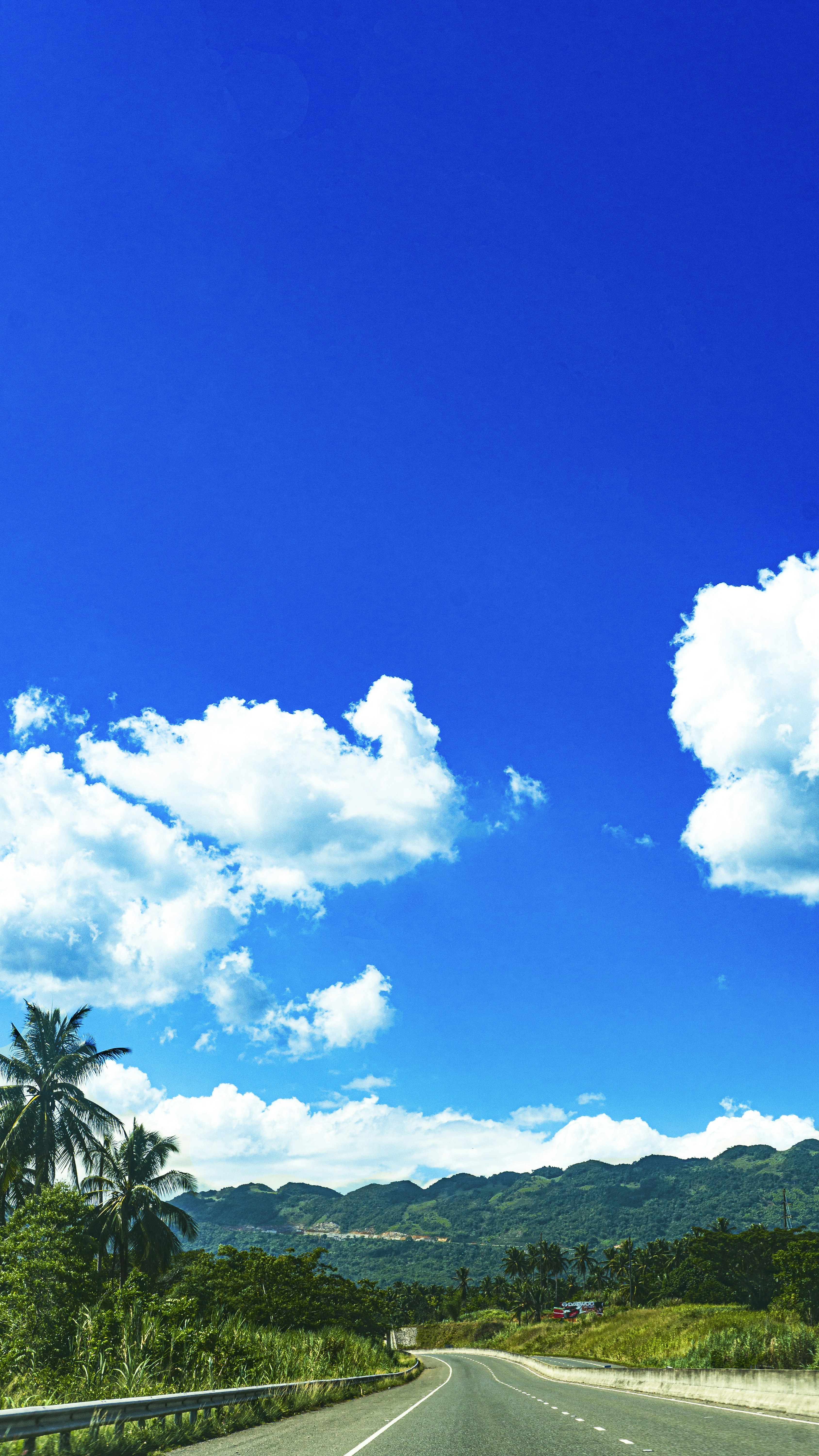 Curving road lined with lush greenery under a vivid blue sky dotted with fluffy clouds.