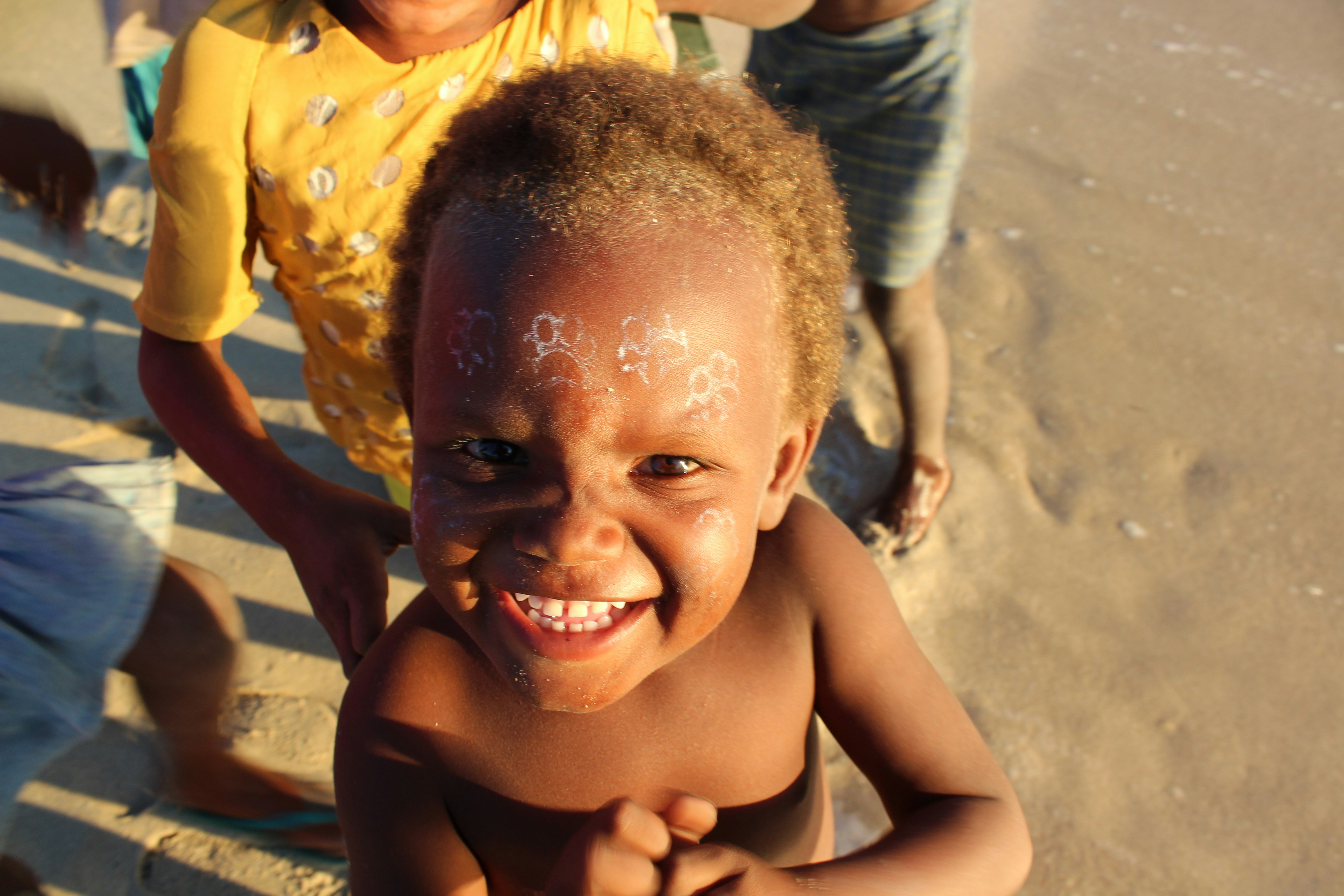 Child smiling brightly on a sandy beach with sunlight casting playful shadows.