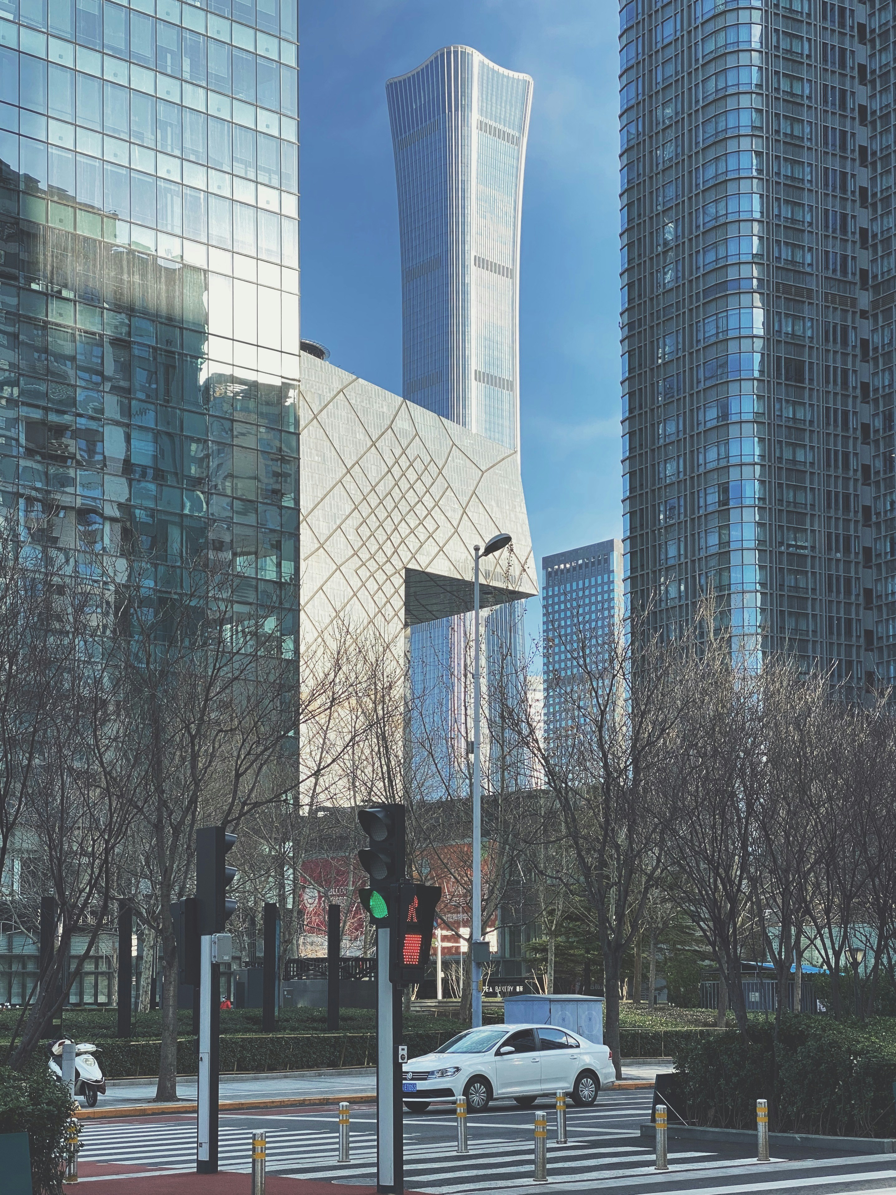 Modern skyscrapers reflect in the glass facade of a unique architectural structure, framed by bare trees and urban elements. 