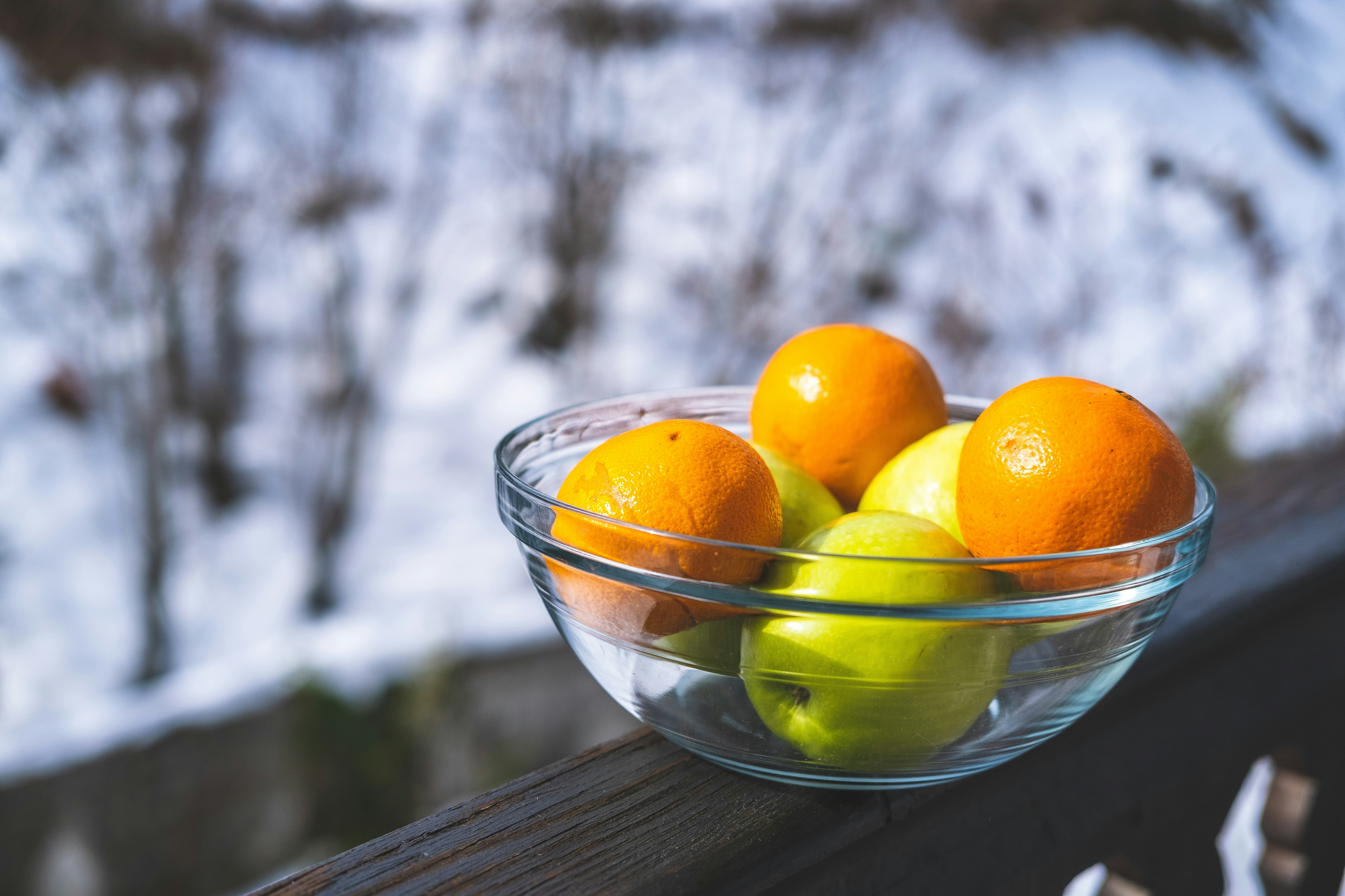 A glass bowl filled with vibrant oranges and green apples sits on a wooden railing, contrasting against a snowy background.