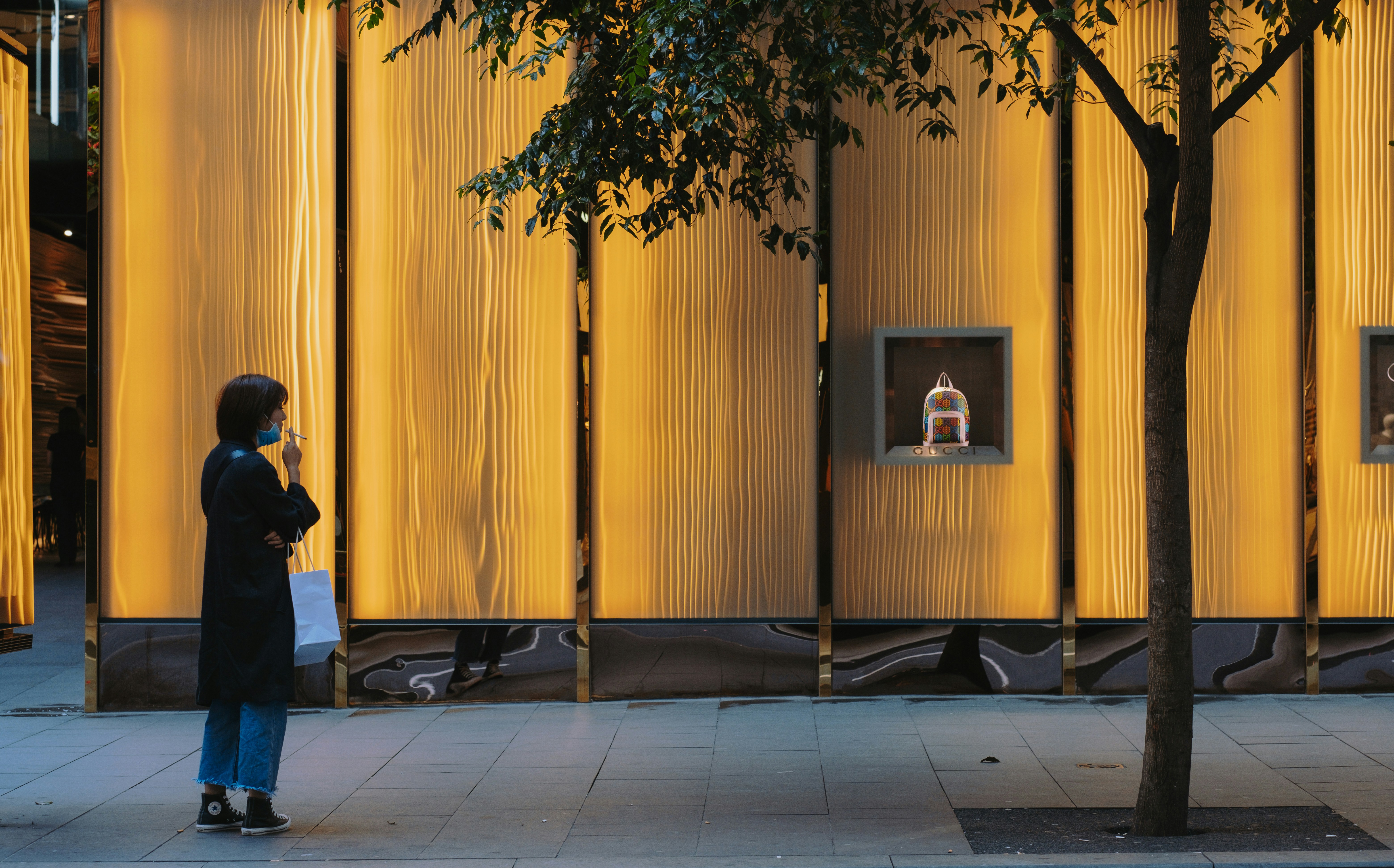 A woman pauses thoughtfully in front of a brightly lit display, framed by textured golden panels and a nearby tree, creating a contrast of nature and modern design.