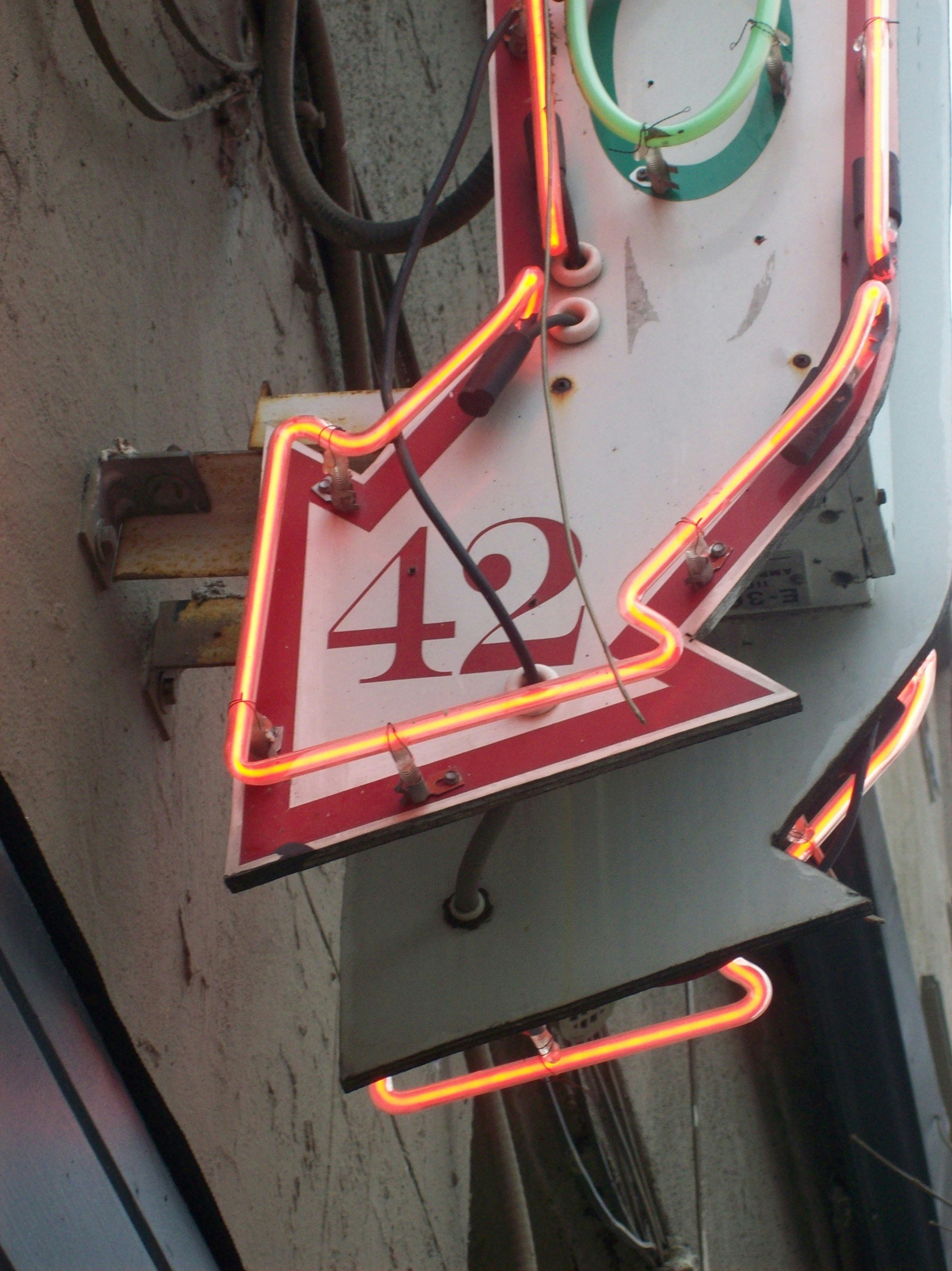 Neon sign displaying the number 42 mounted on a weathered wall, with exposed wiring and looping tubing. The warm orange glow contrasts with the rough surface and metal brackets.