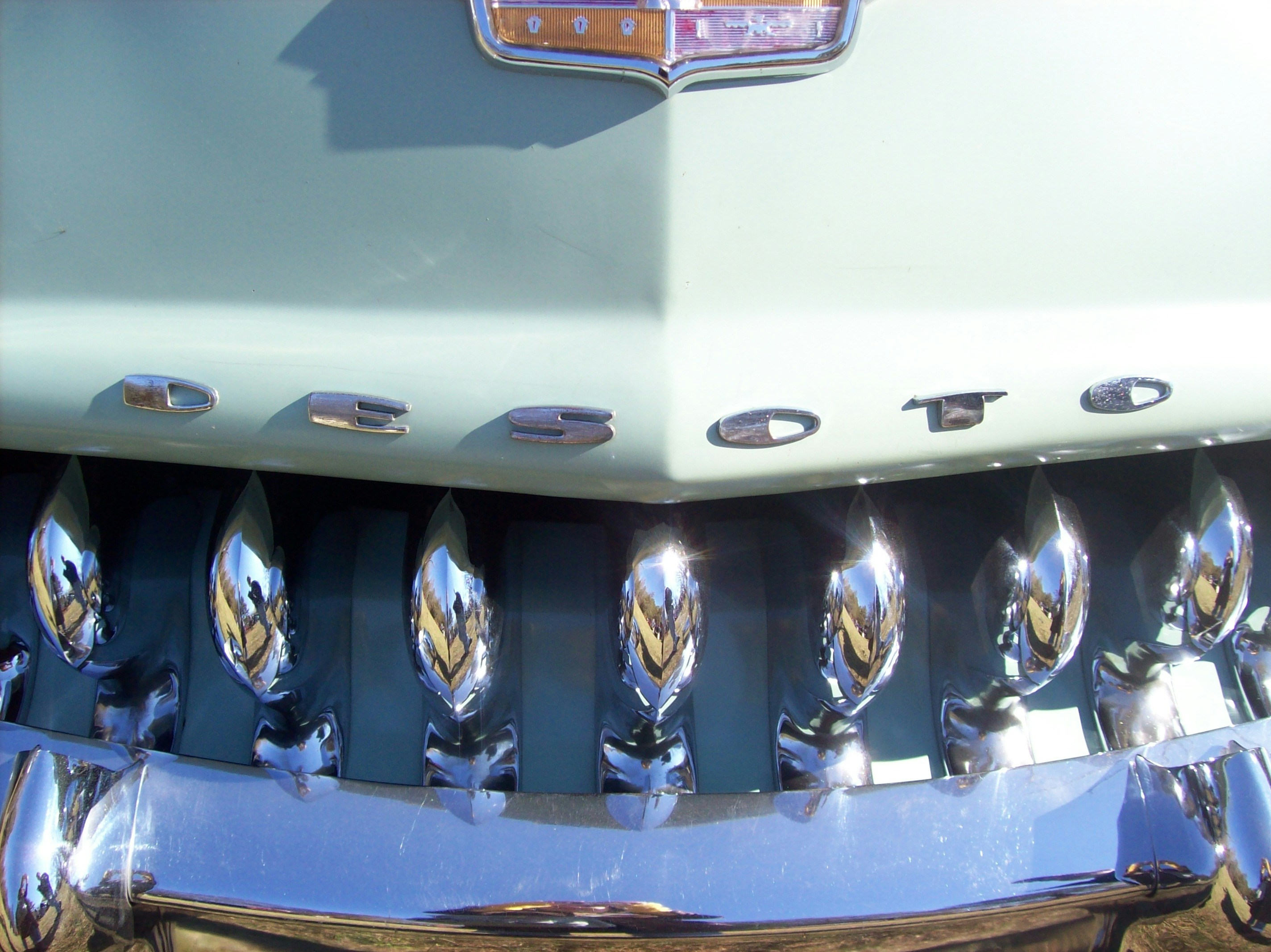 Close-up of a pastel-green vintage car front, highlighting a chrome grille with bullet-like vertical bars and the emblem above. Reflections shimmer across the chrome and bumper.