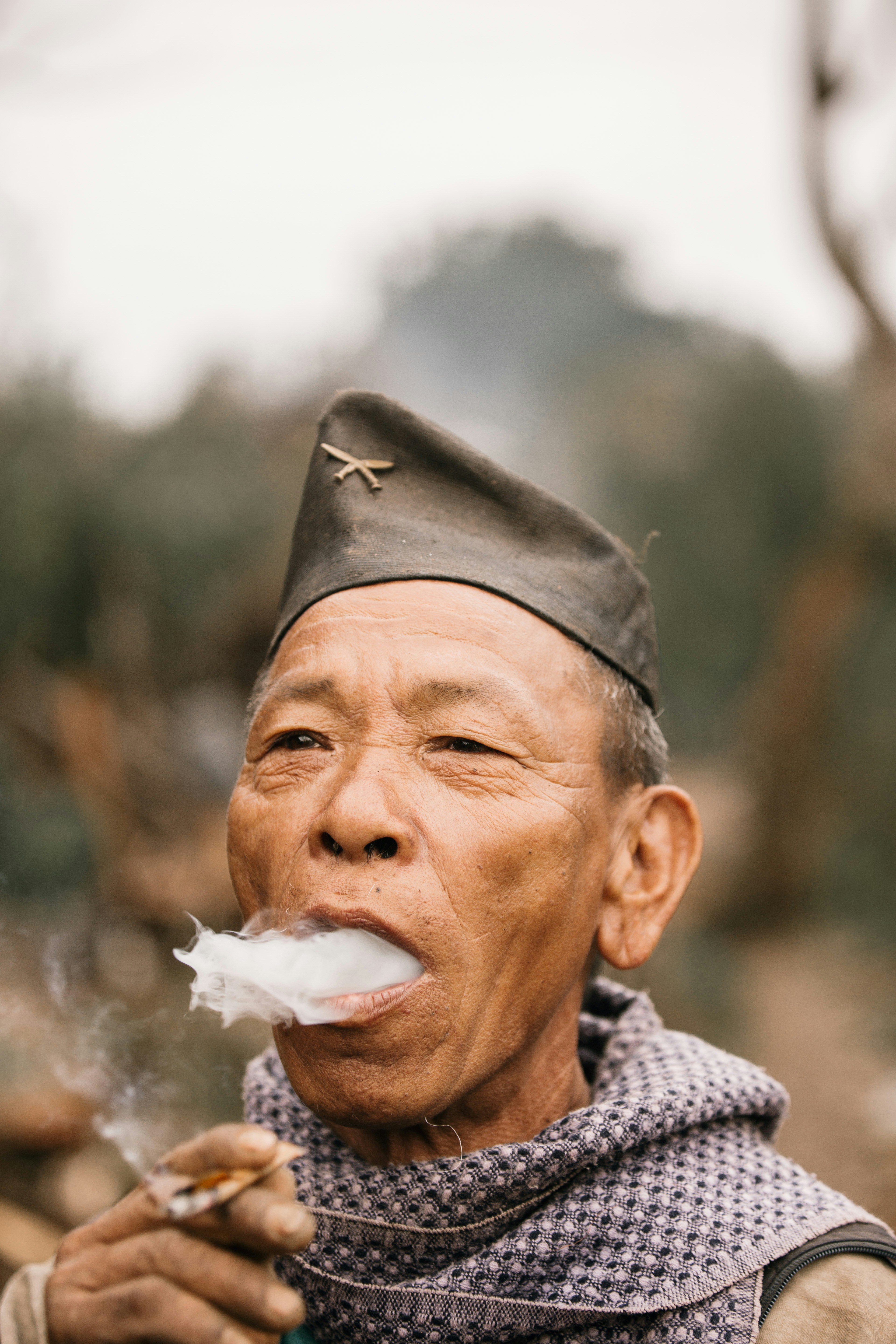 Elderly man exhaling smoke while wearing a traditional hat and scarf, surrounded by a blurred natural background.