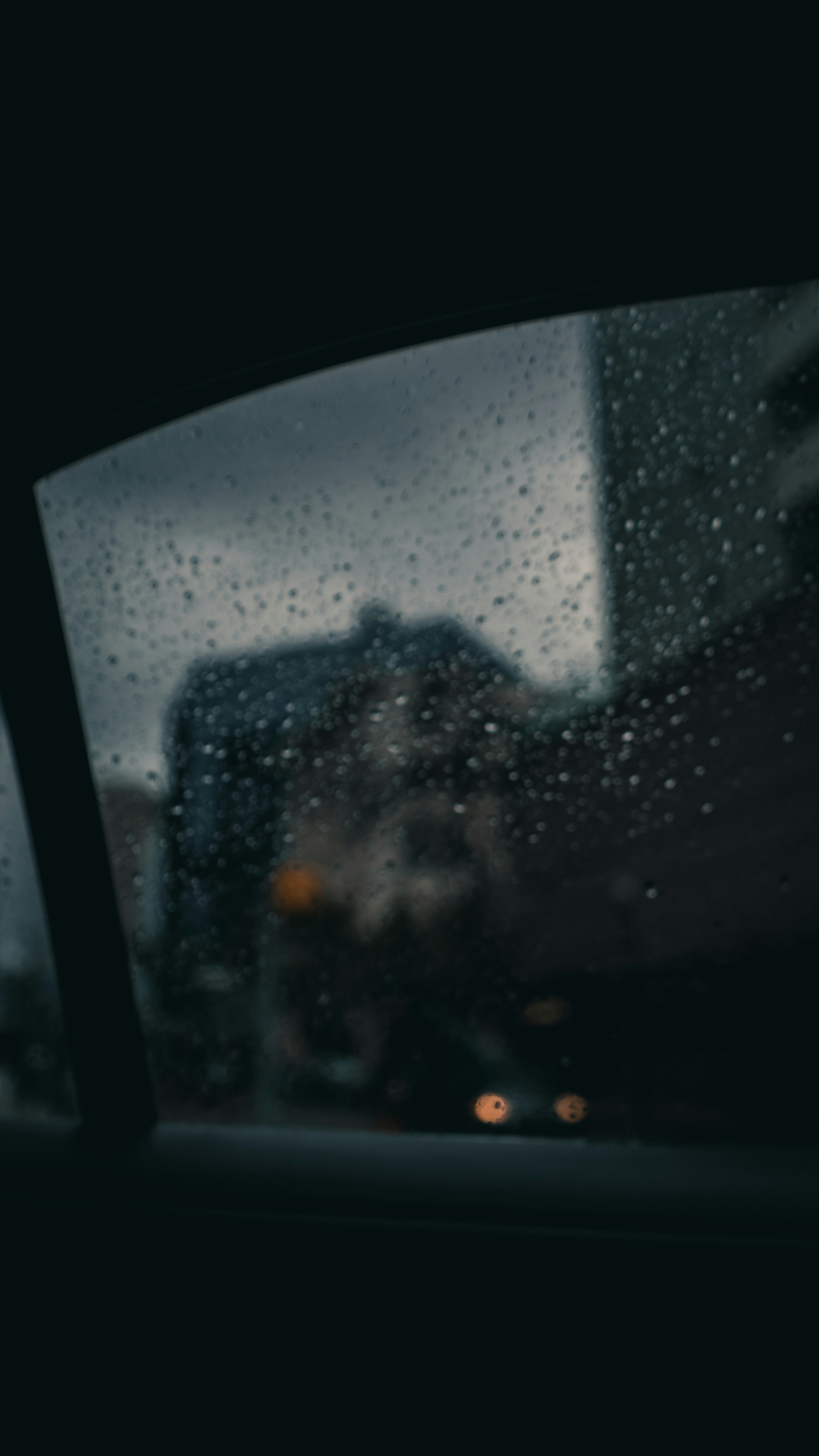 Raindrops cling to a car window, blurring the view of a city street illuminated by soft lights. The atmosphere evokes a sense of introspection and urban solitude.