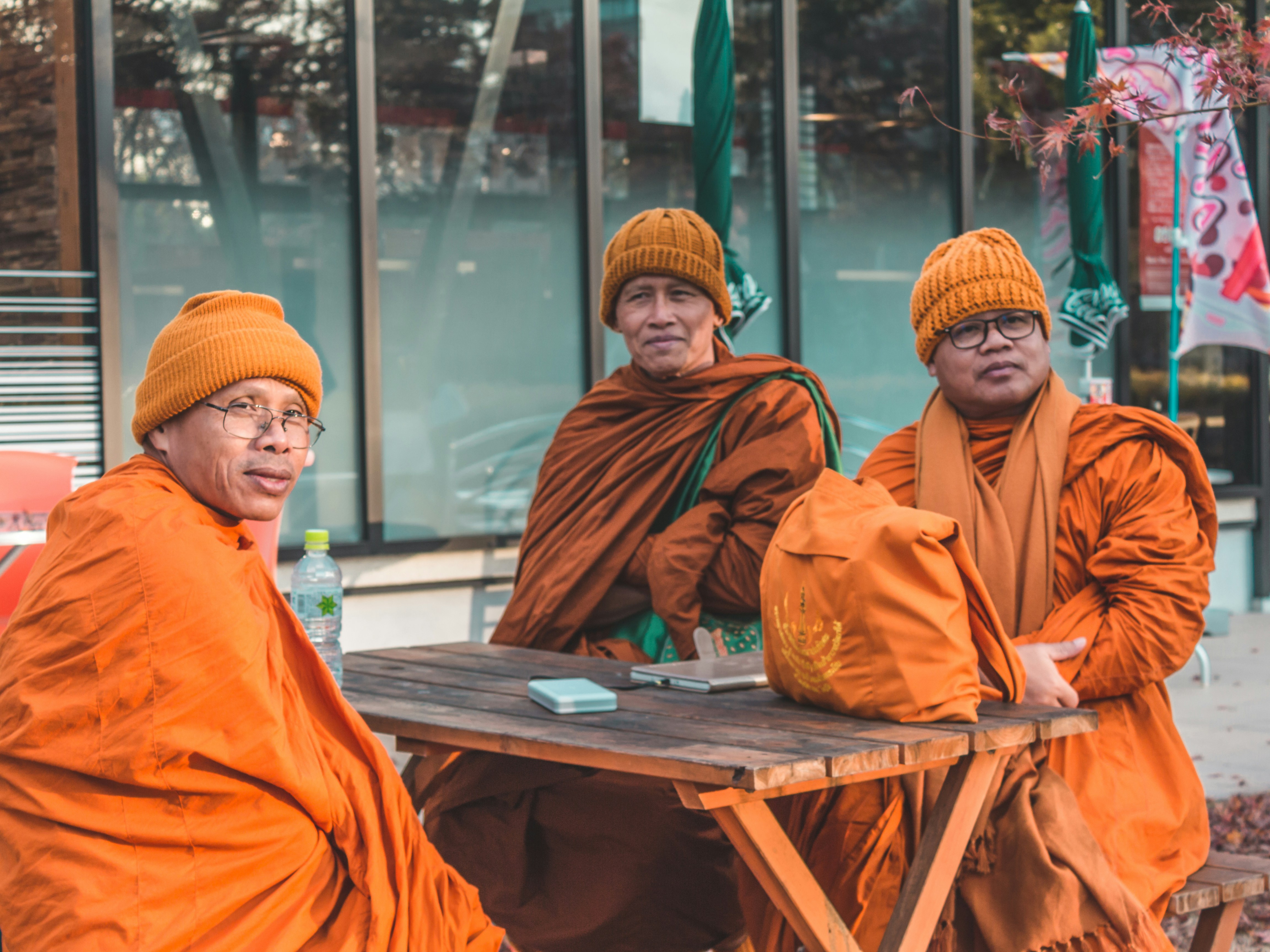 Three monks in orange robes seated at a wooden table outdoors.