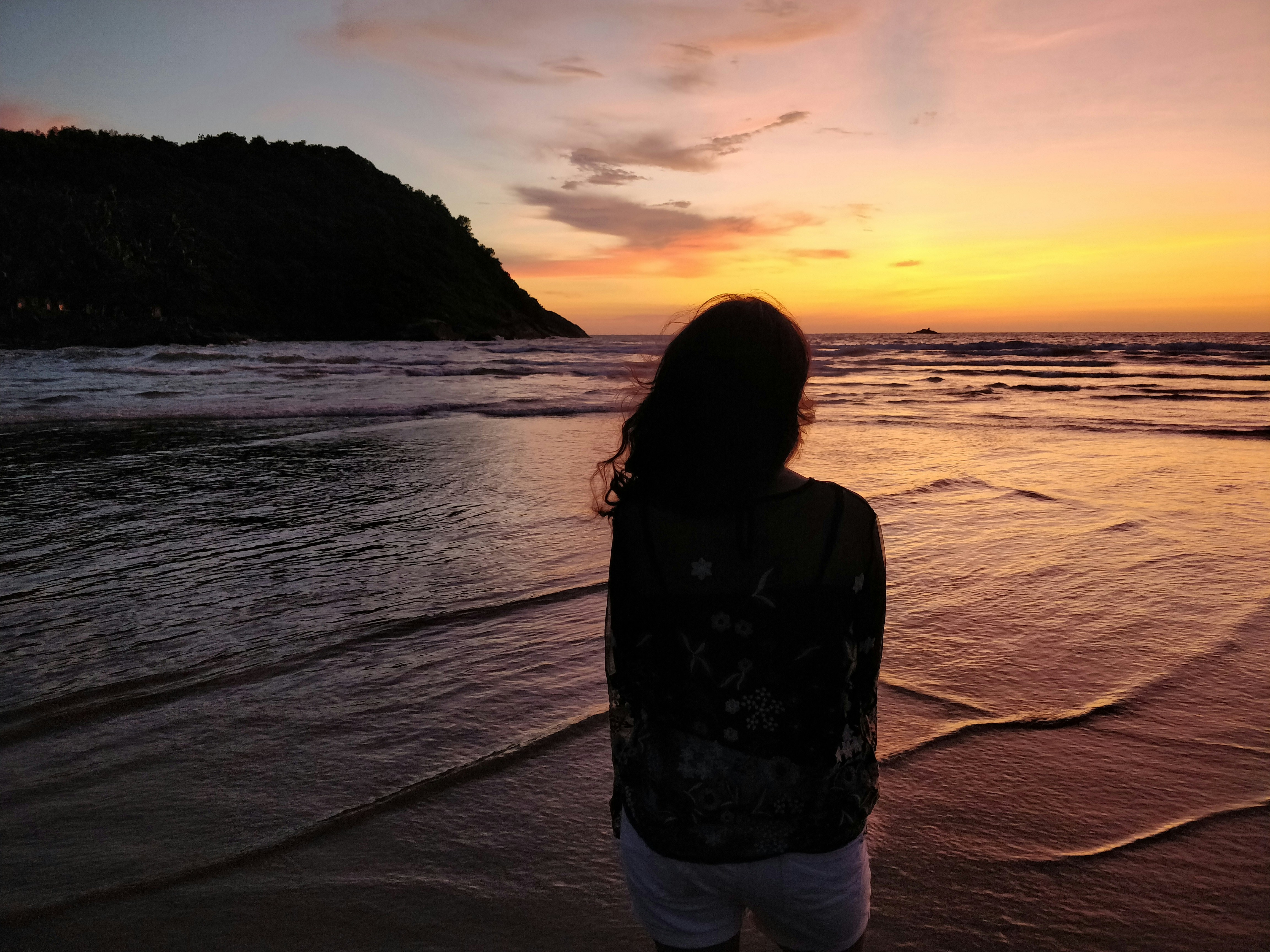 Silhouette of a person standing on a beach during a vibrant sunset, with waves gently washing over the sand.