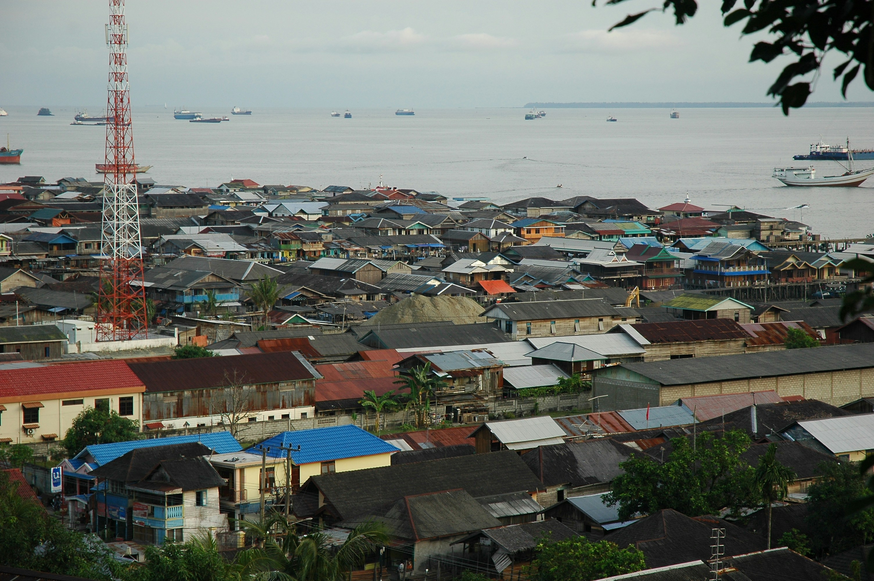 Seaside community with densely packed rooftops and a distant view of ships on calm waters.