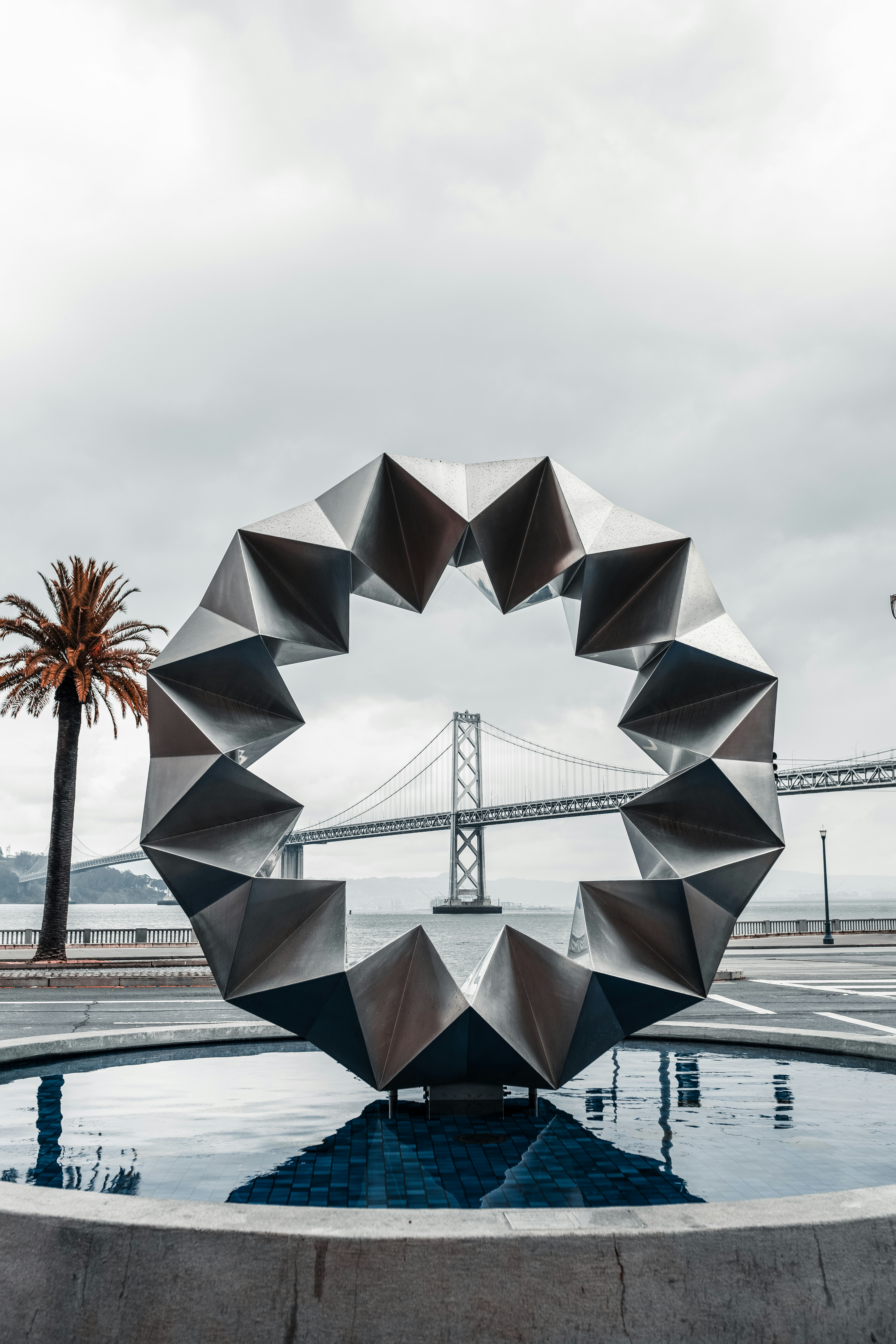 Sculptural fountain featuring a geometric design, framed by a palm tree and the Bay Bridge in the background. The scene captures a blend of urban art and nature.