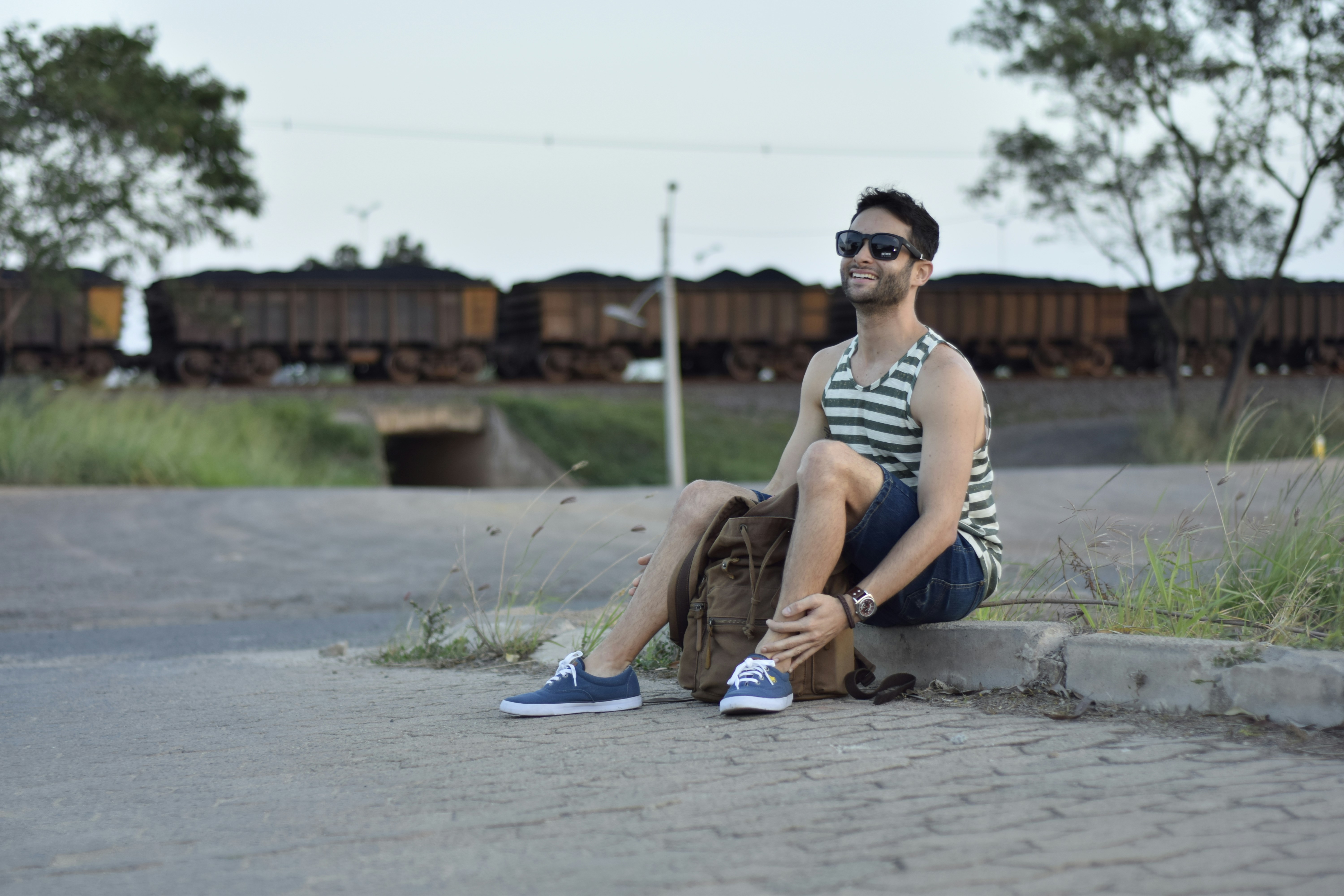 Person in a striped tank top sitting on a curb near passing train cars, with trees in the background.