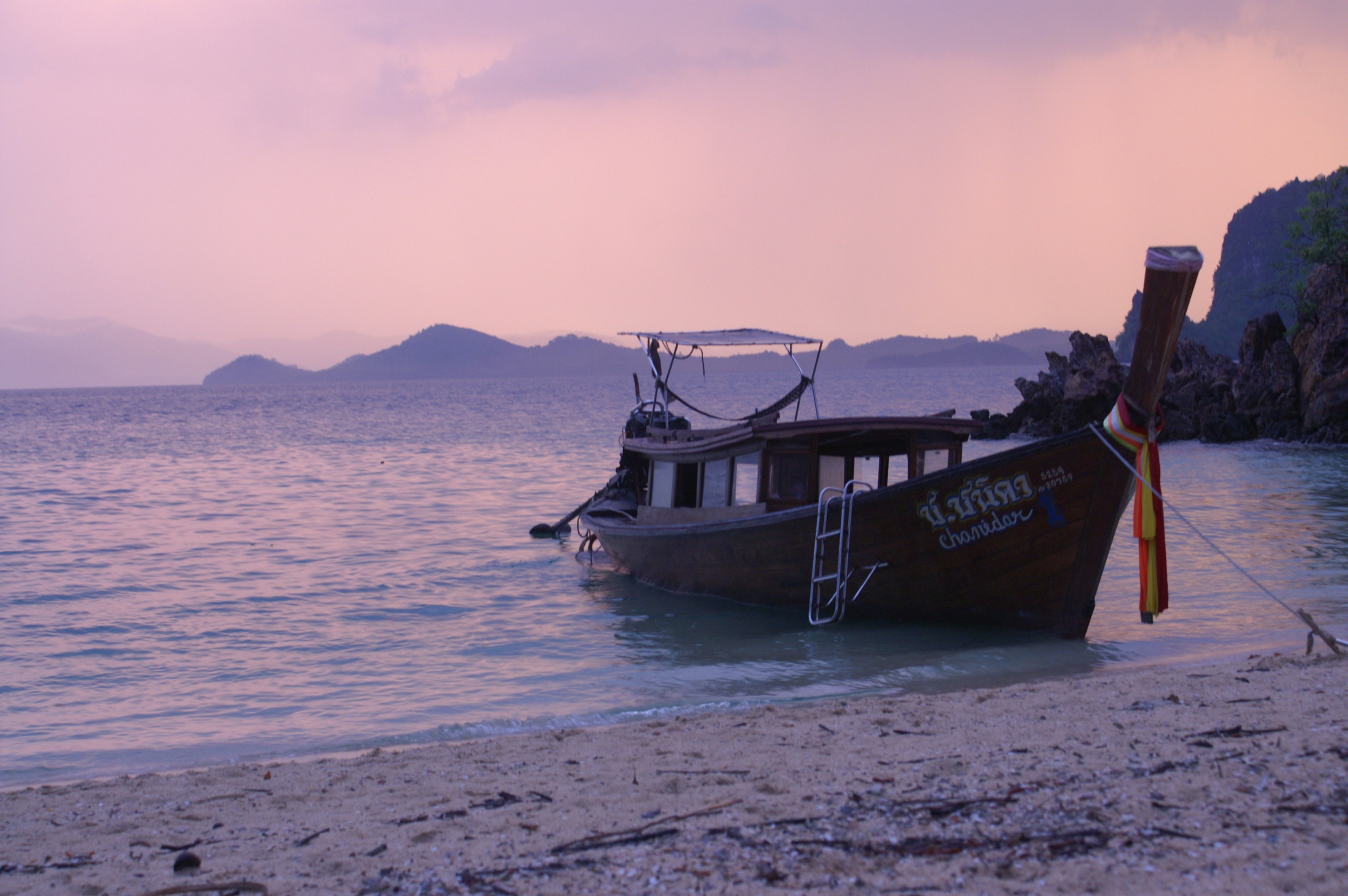 Wooden boat resting on a sandy beach at sunset with distant hills under a purple sky.