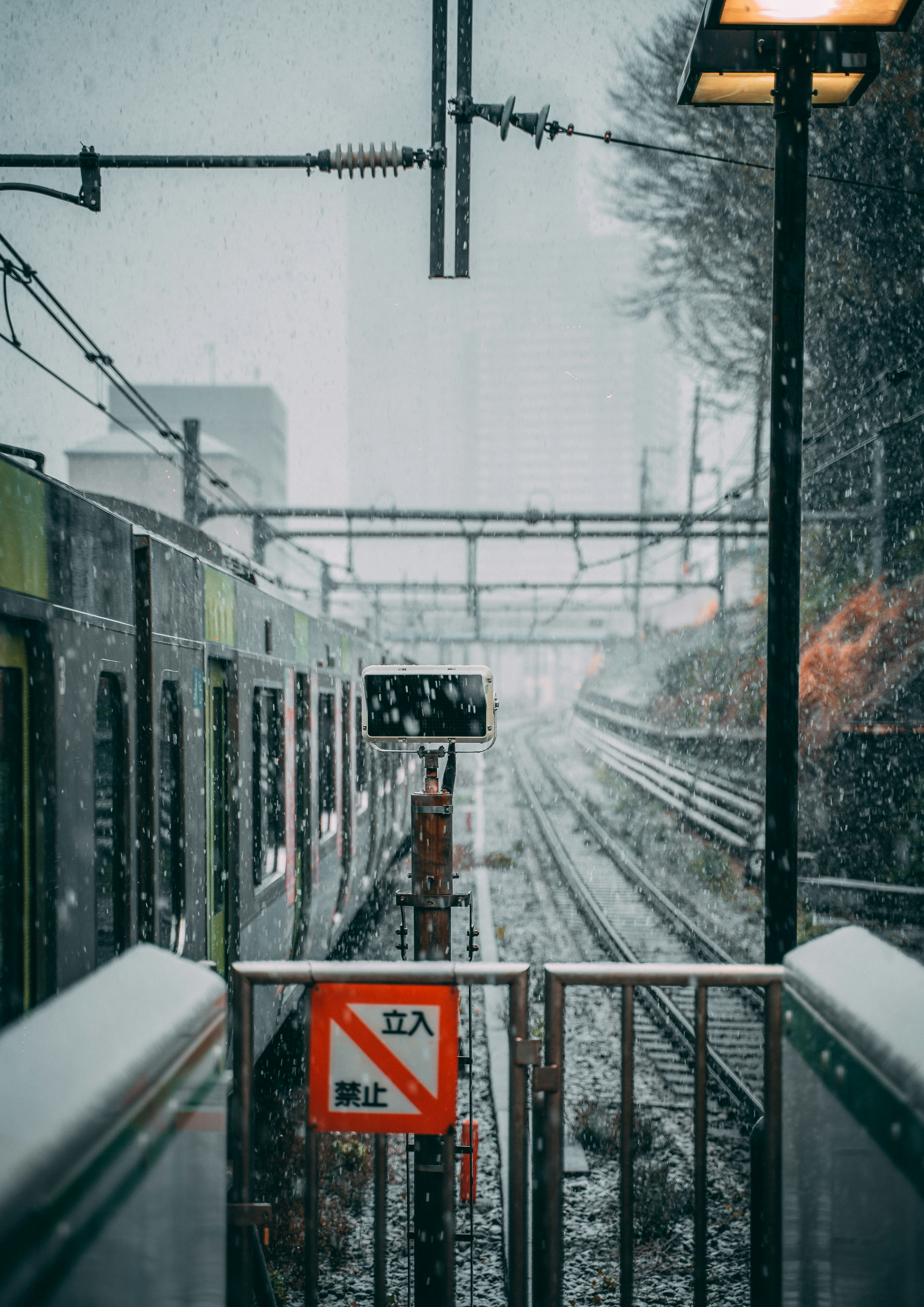 Snowfall blankets a quiet train station, with a train waiting beside a signaling post. The scene evokes a sense of stillness amidst the flurry.