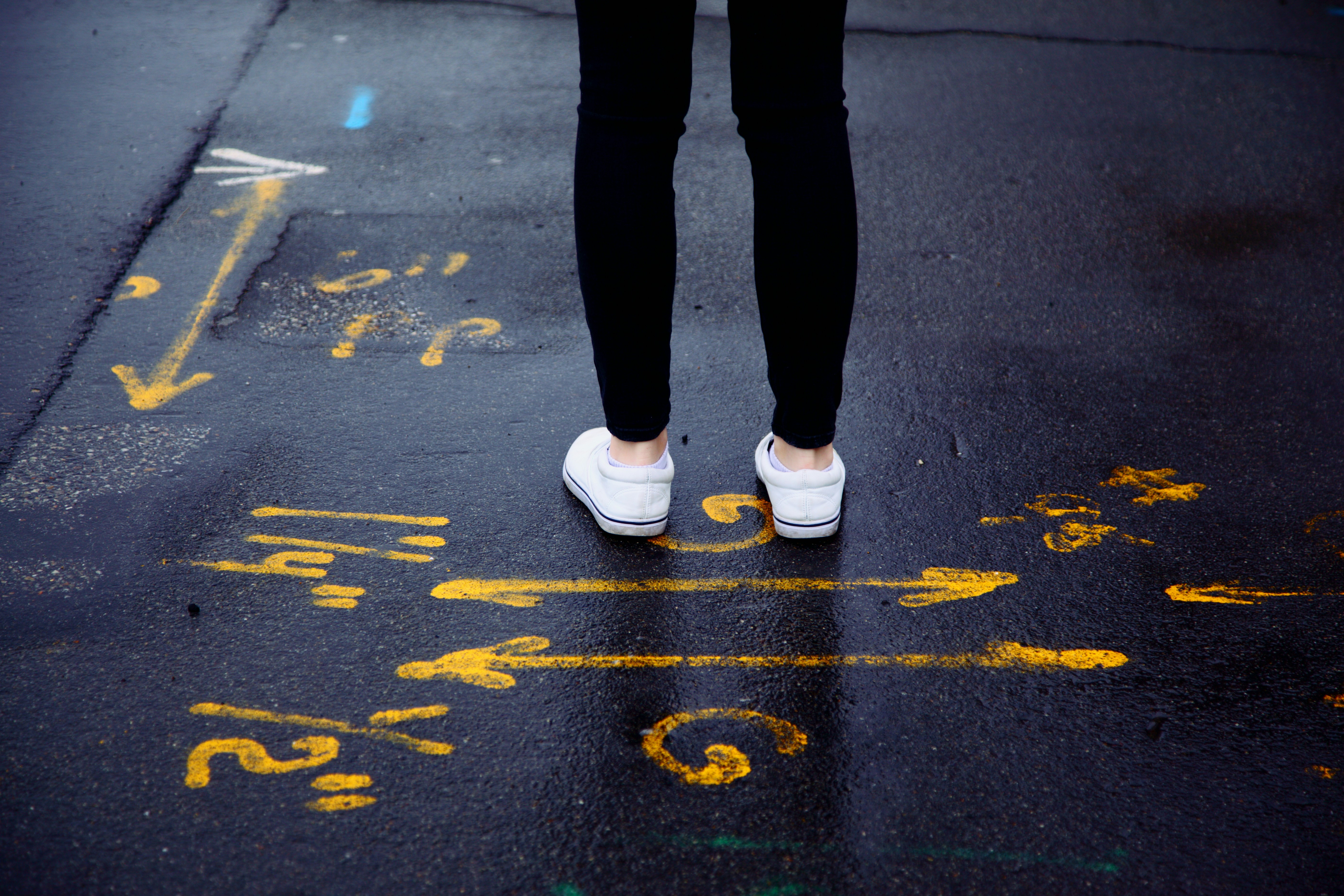 Person in black pants and white sneakers standing on asphalt marked with yellow construction lines.