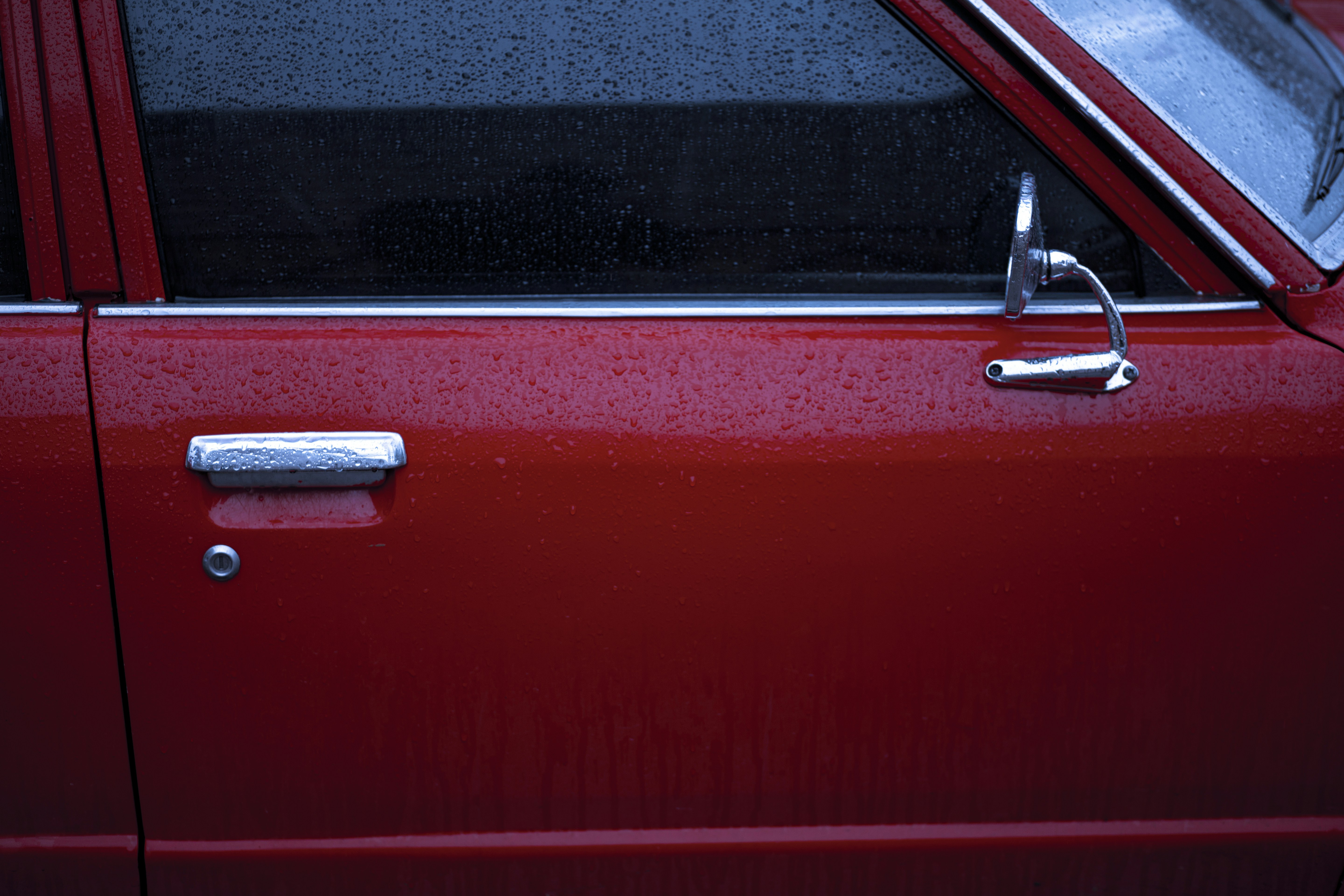 Close-up of a red car door with raindrops glistening under overcast skies.